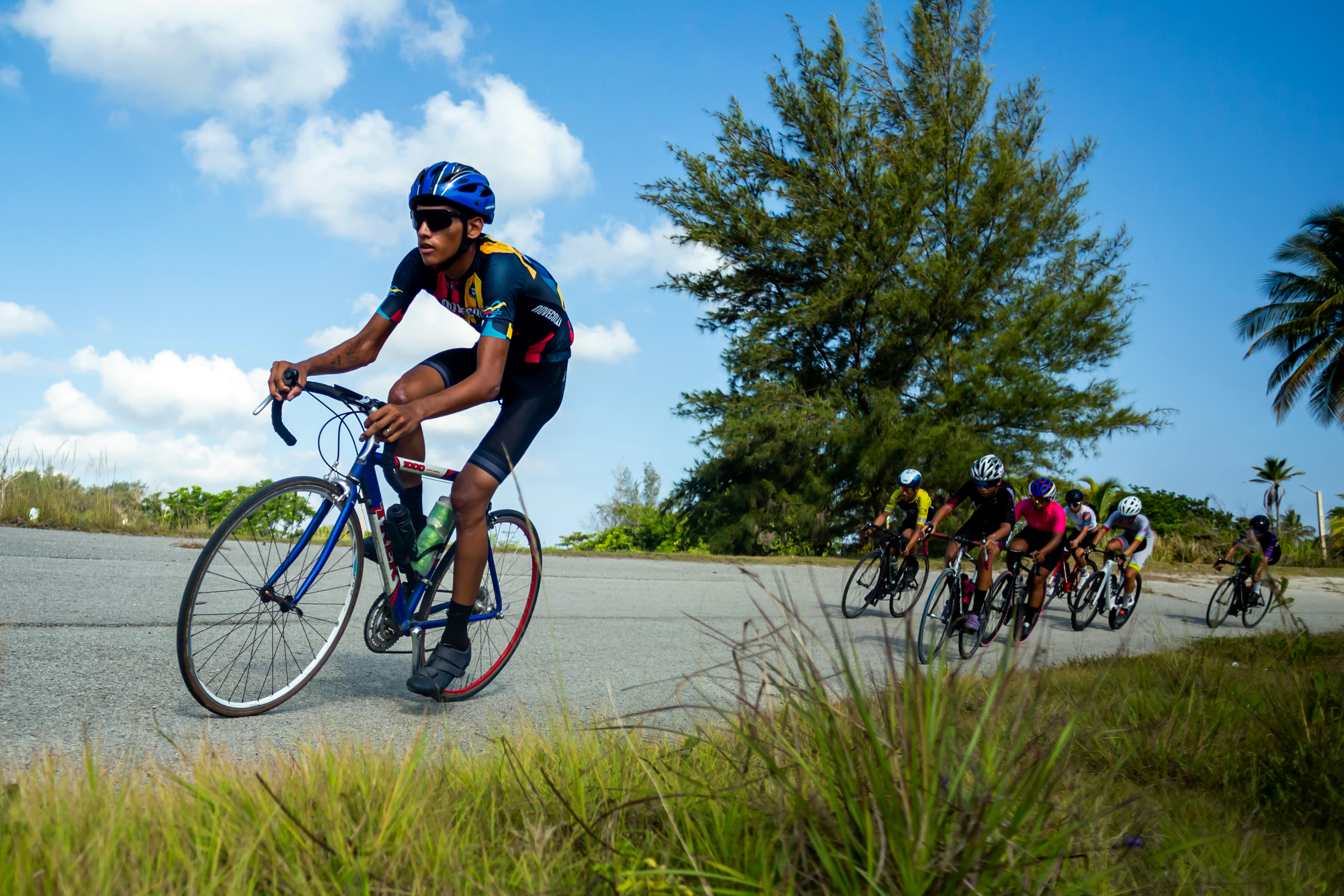 A group of cyclists racing on a sunlit road, with the lead rider in a blue helmet foregrounded while teammates trail behind; the scene captures motion and bright outdoor colors. (Note: this is a photo, not a painting or illustration.)