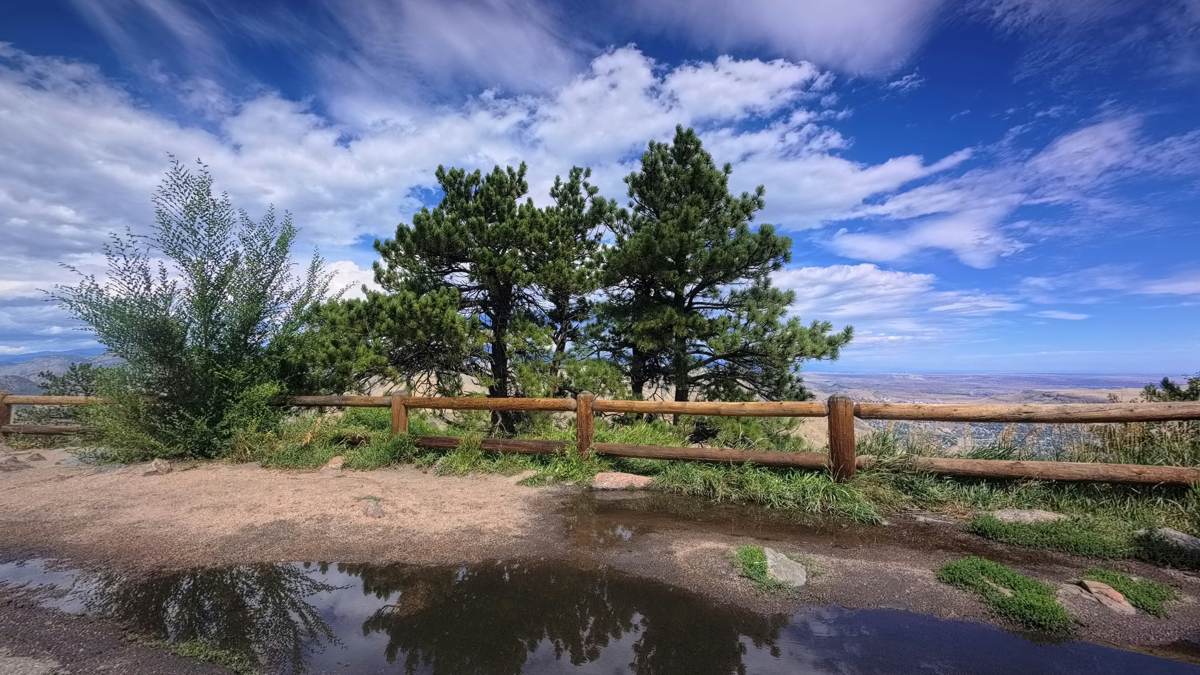 Wide overlook with a wooden railing, pines and shrubs under a bright blue sky; a puddle reflects the scene, hinting at recent rain.
