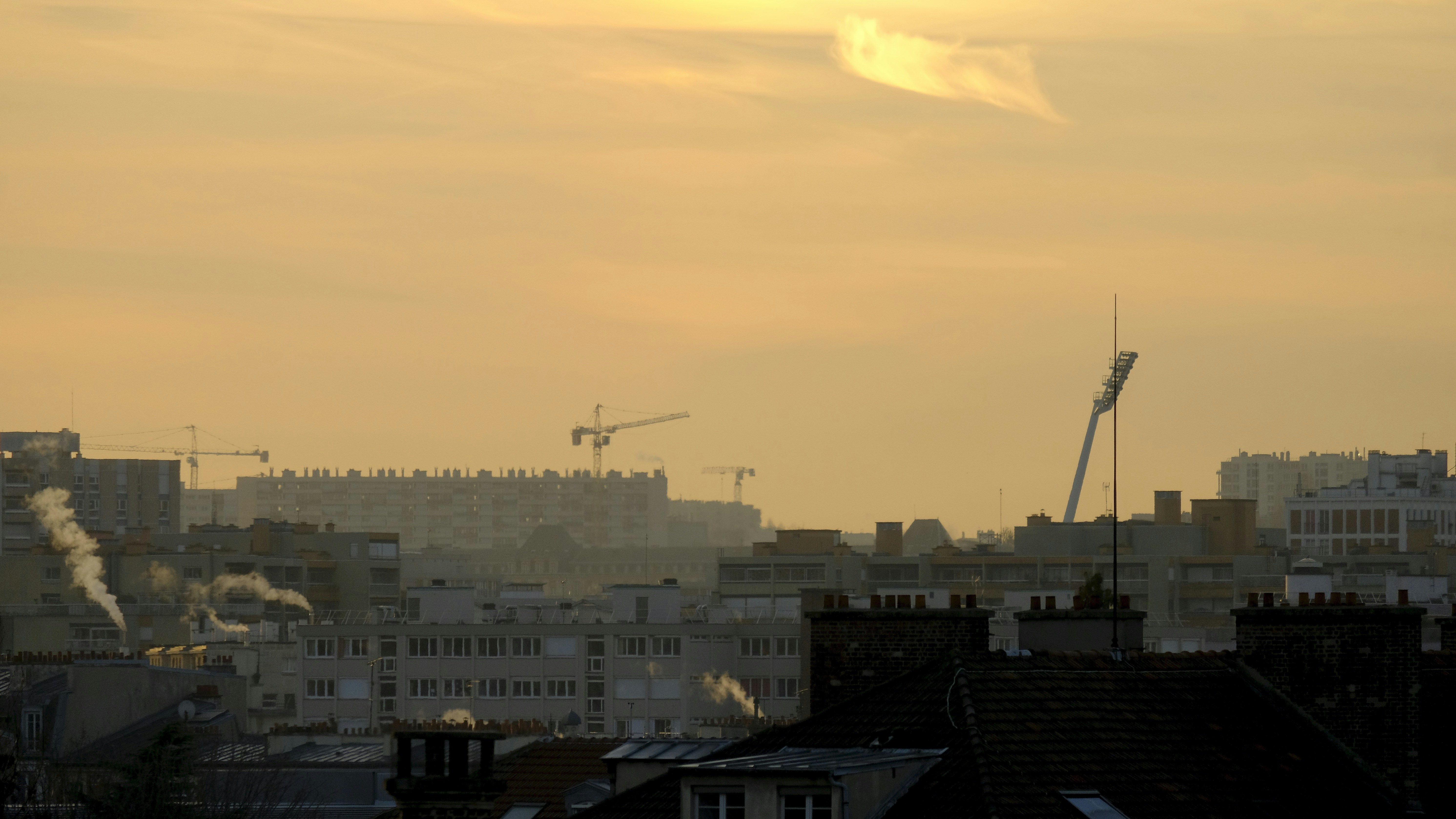 A hazy cityscape at sunset, silhouettes of buildings and chimneys across a calm horizon, with warm tones and distant cranes hinting at ongoing development.