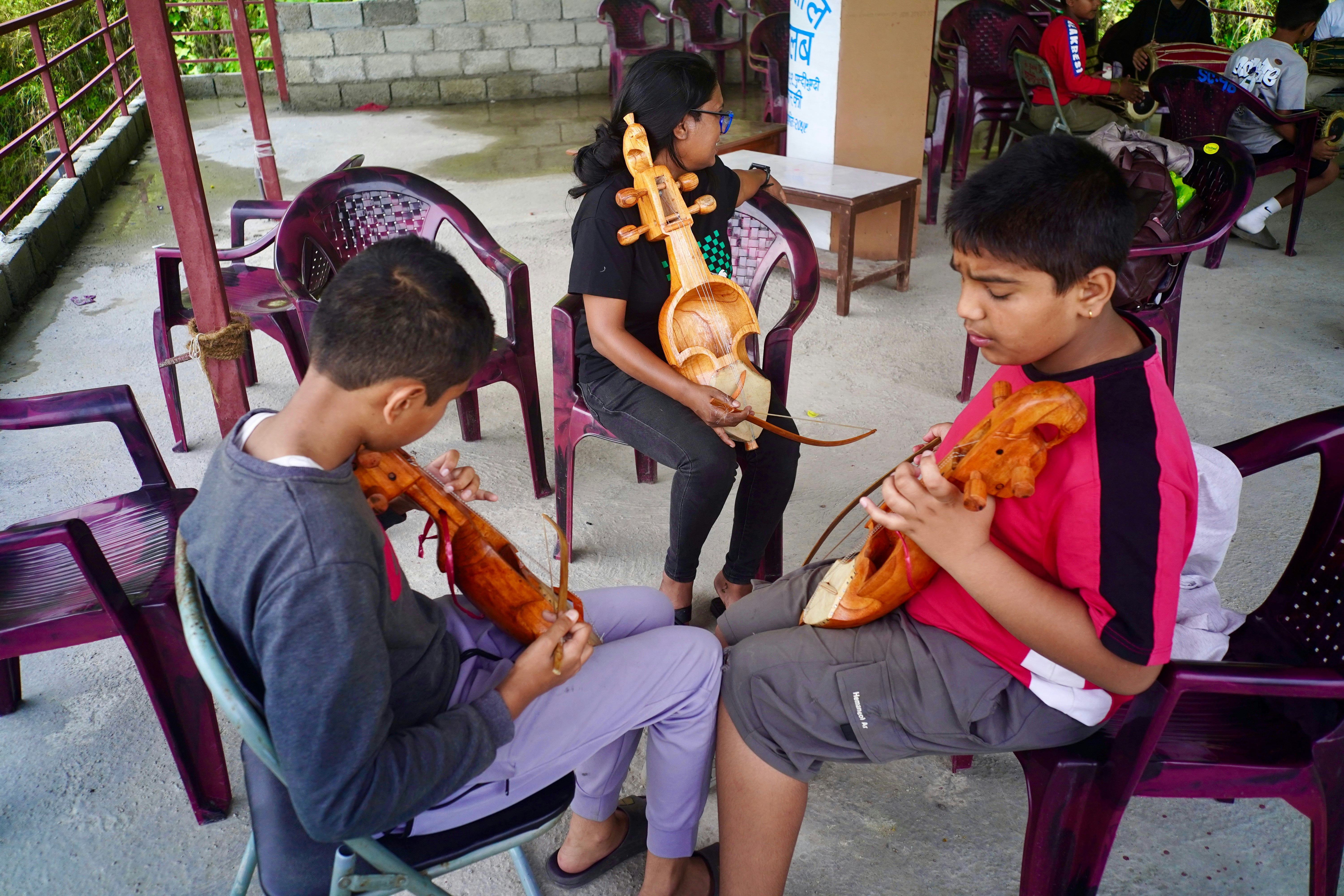 Three children sit on chairs in an outdoor covered area, each playing a small violin; one in the foreground left, one in the foreground right, and another seated behind them with a violin. The scene resembles a casual, informal practice or jam session.