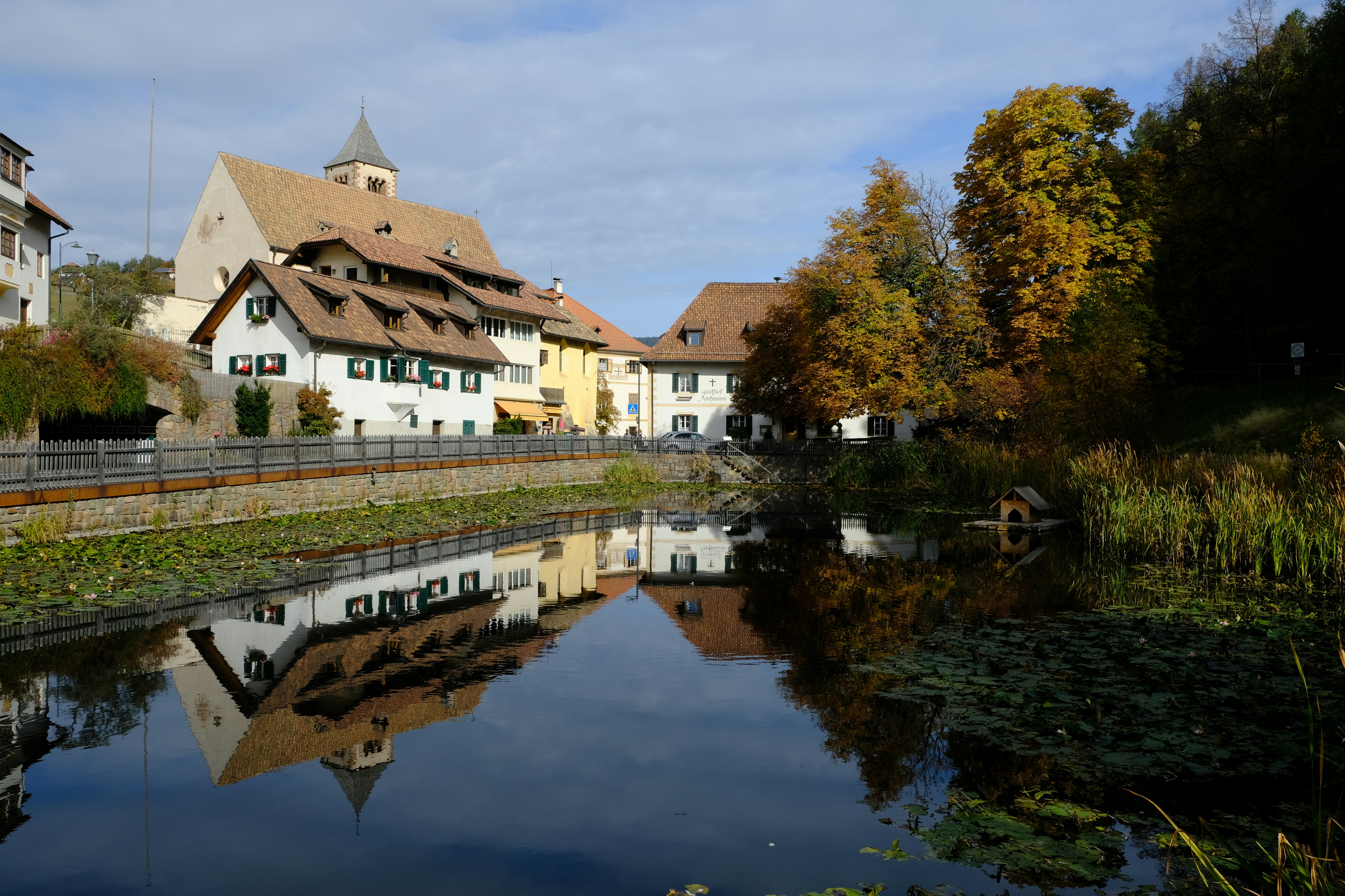 Colorful village buildings line a calm canal, perfectly reflected in the water with autumn trees on the right. A tranquil scene showing architecture and natural scenery together.