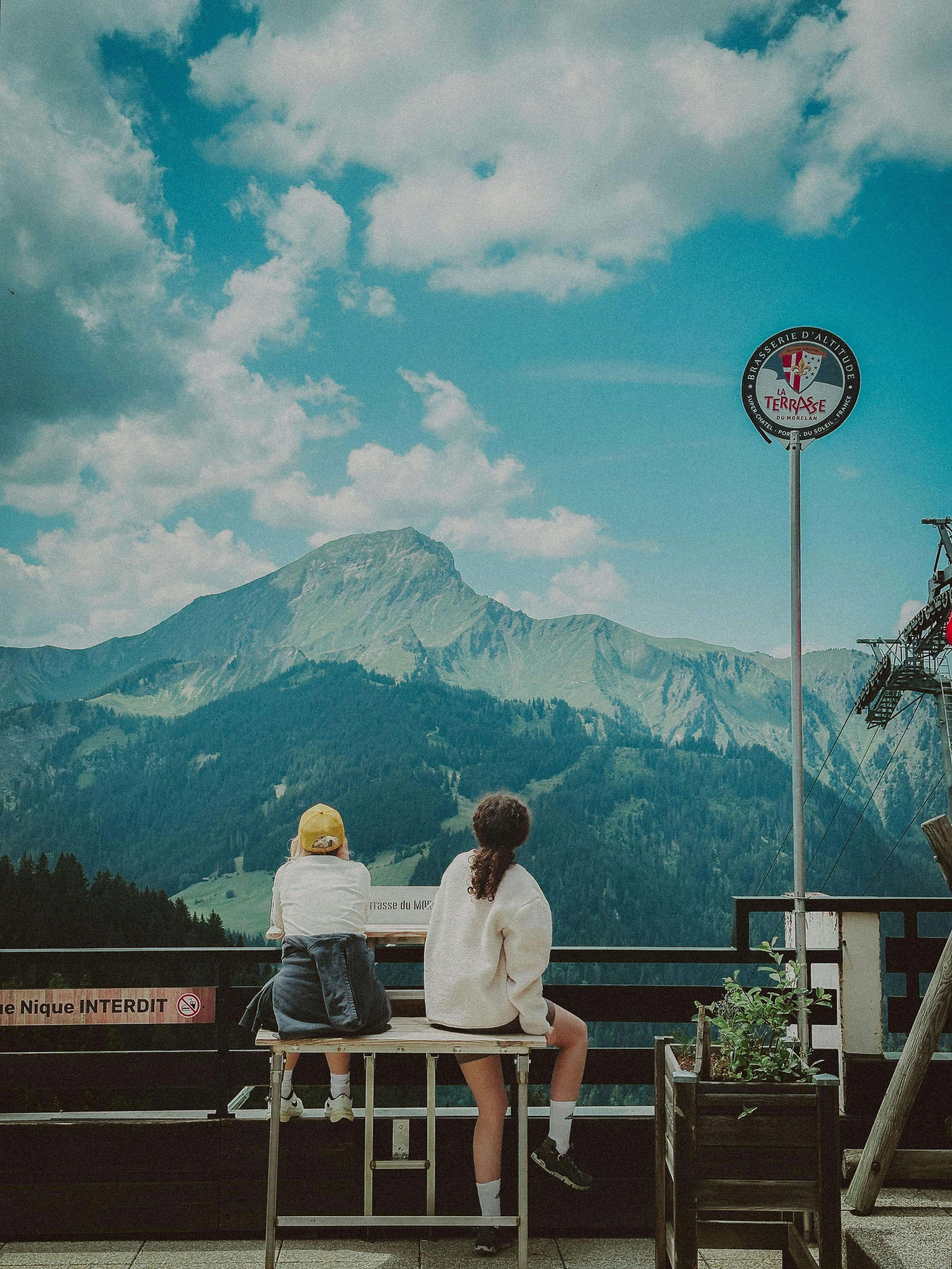 Two friends sit on a railing at a scenic mountain overlook, with blue sky and distant peaks in the background.