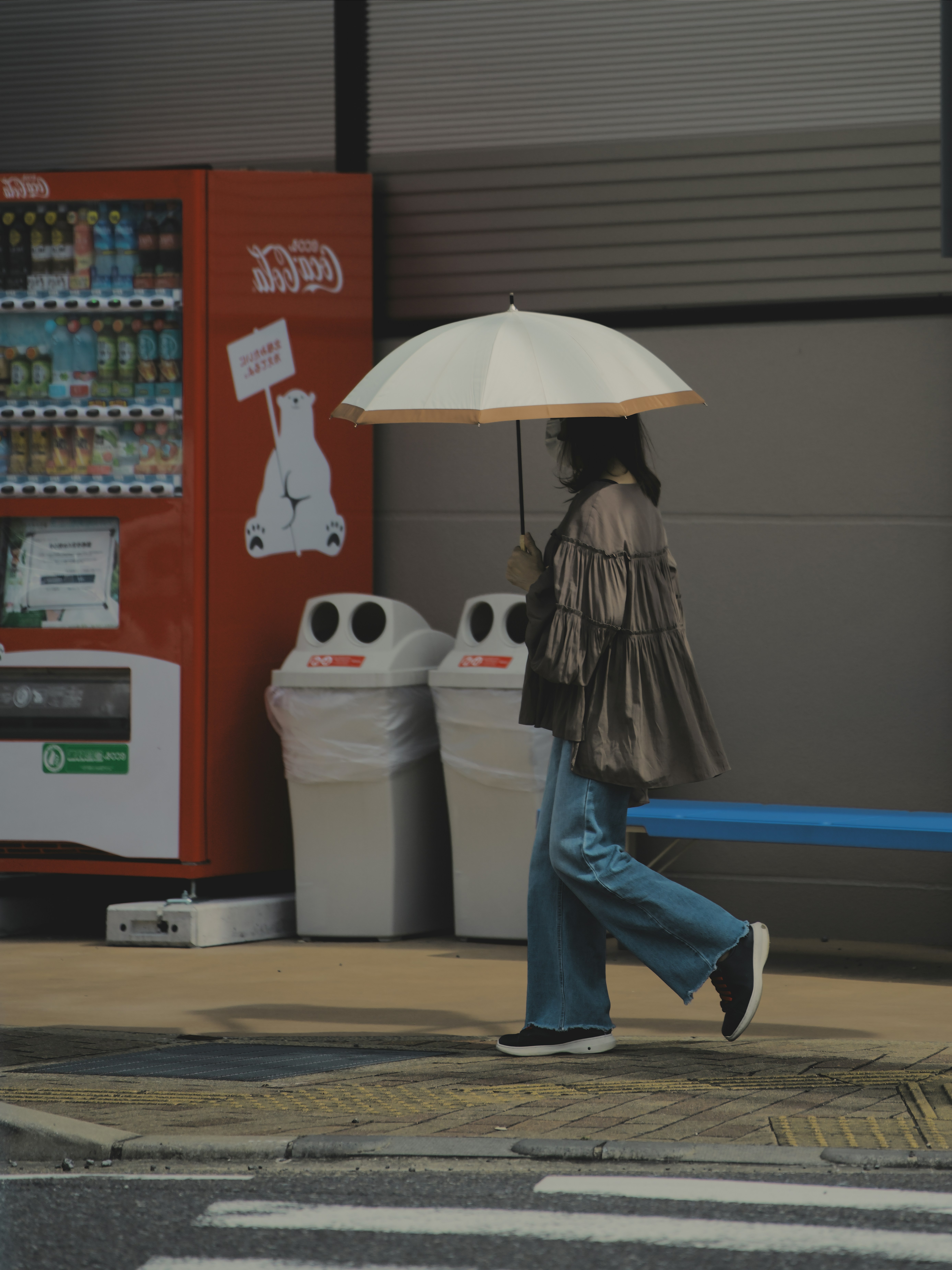 A person walks with an umbrella past a red vending machine and two white trash bins beside a gray wall; a blue bench lines the scene.