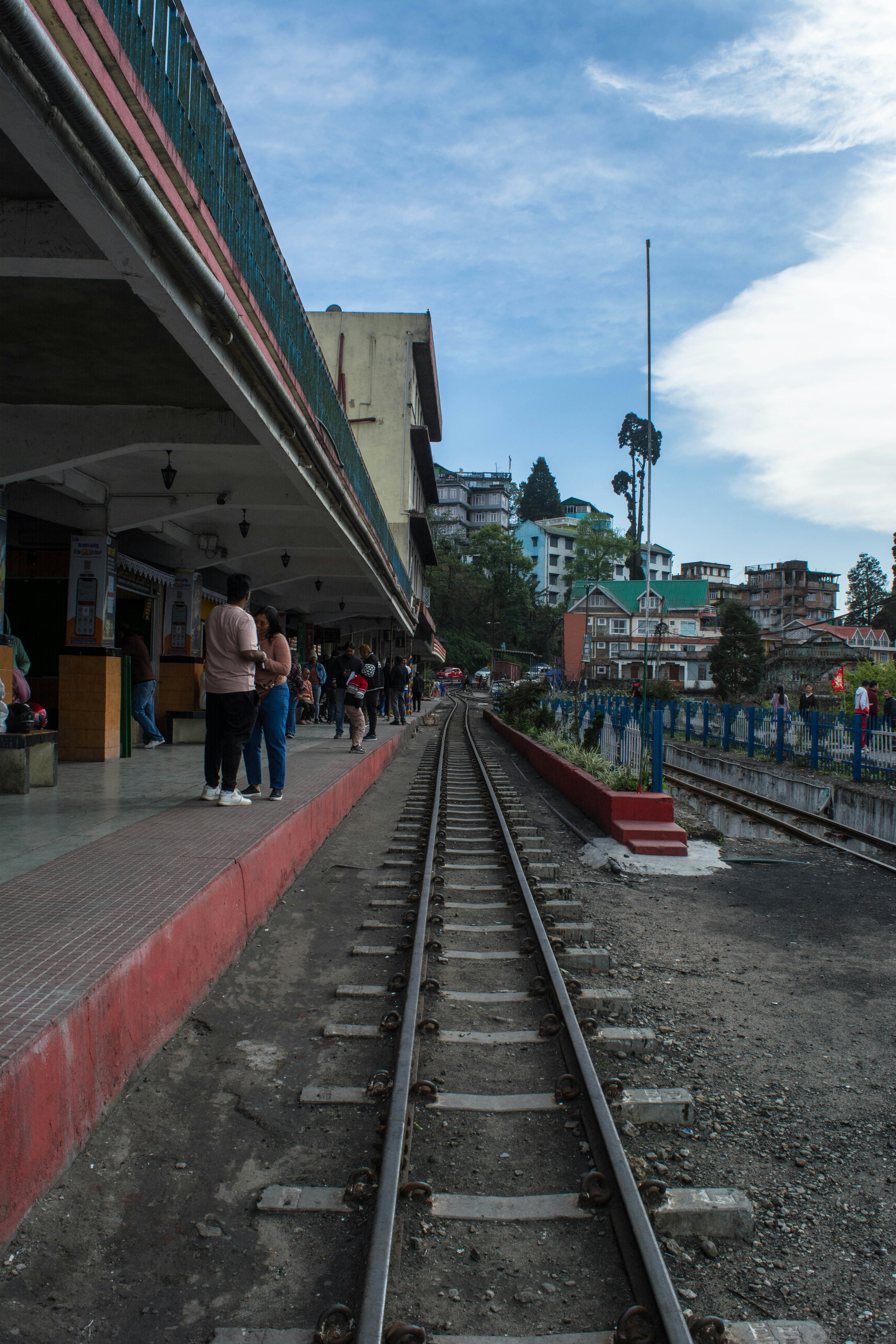People gather along a railway platform beside a fading red curb, with tracks leading into a distant town under a bright skyline.