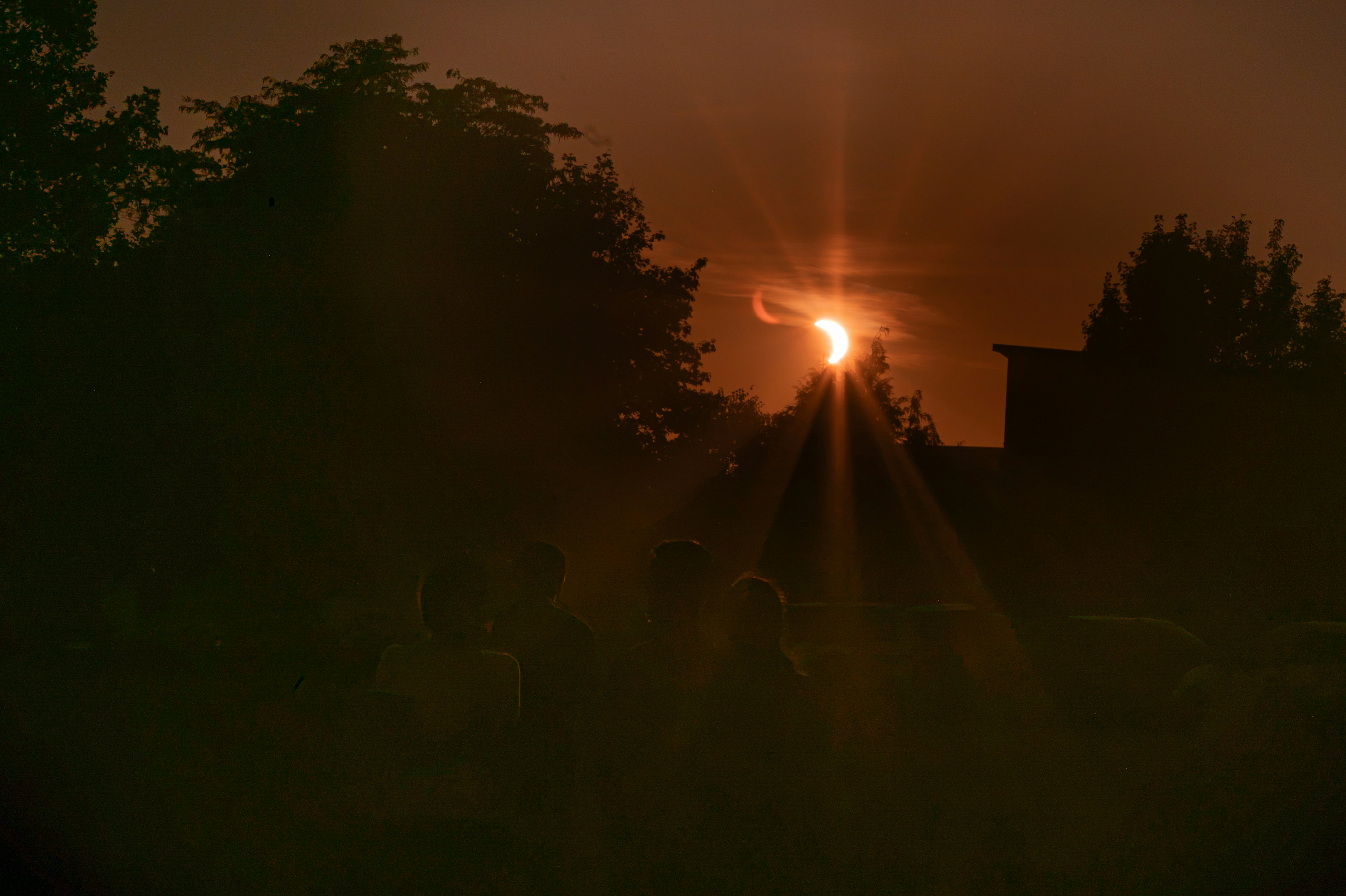 Low-light sunset scene with a bright sunburst behind silhouettes of people and trees; the composition emphasizes a distant horizon and atmospheric haze.