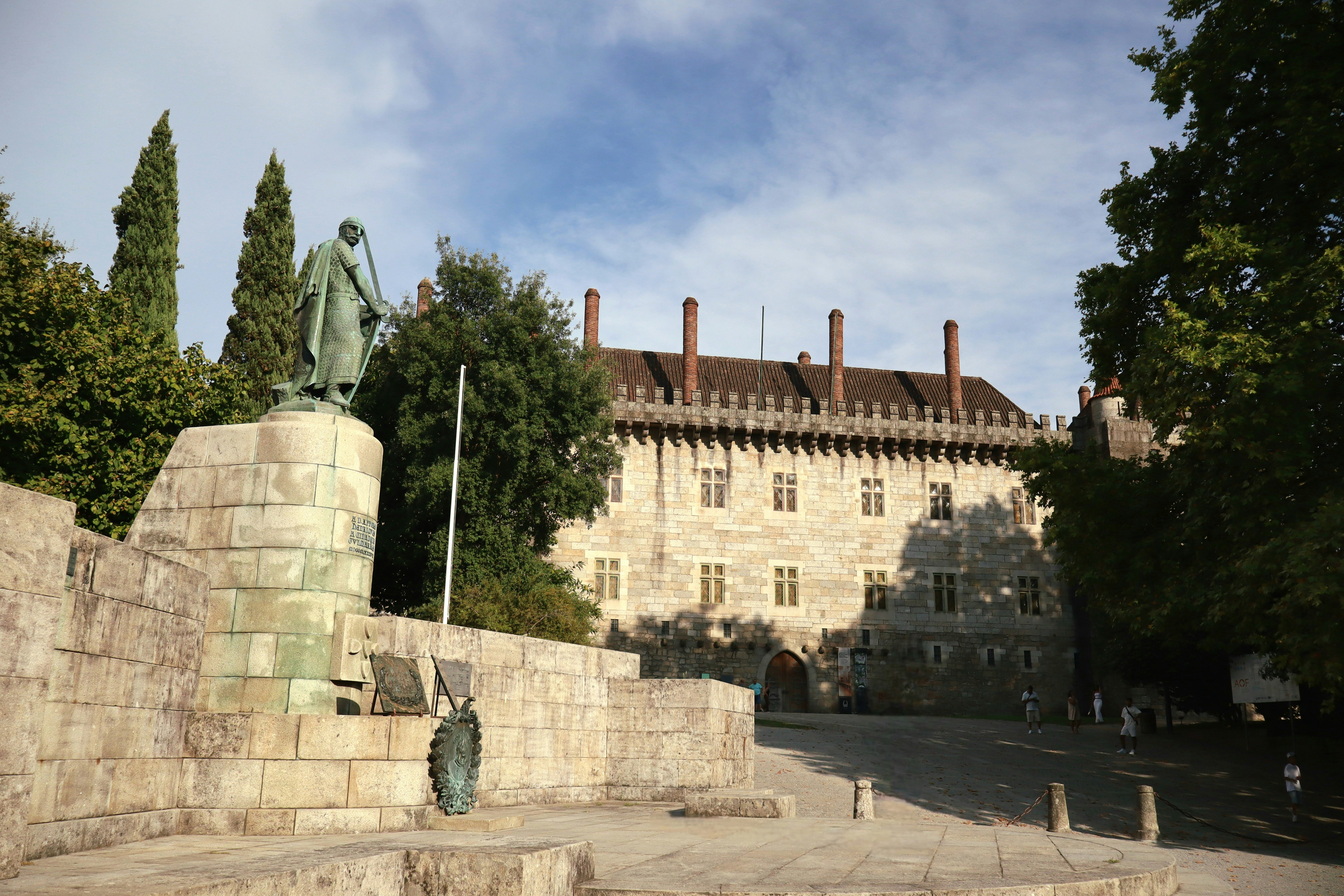 Sunlit stone plaza with a statue on a pedestal in the foreground and an old fortress facade in the background, conveying a historic urban space.