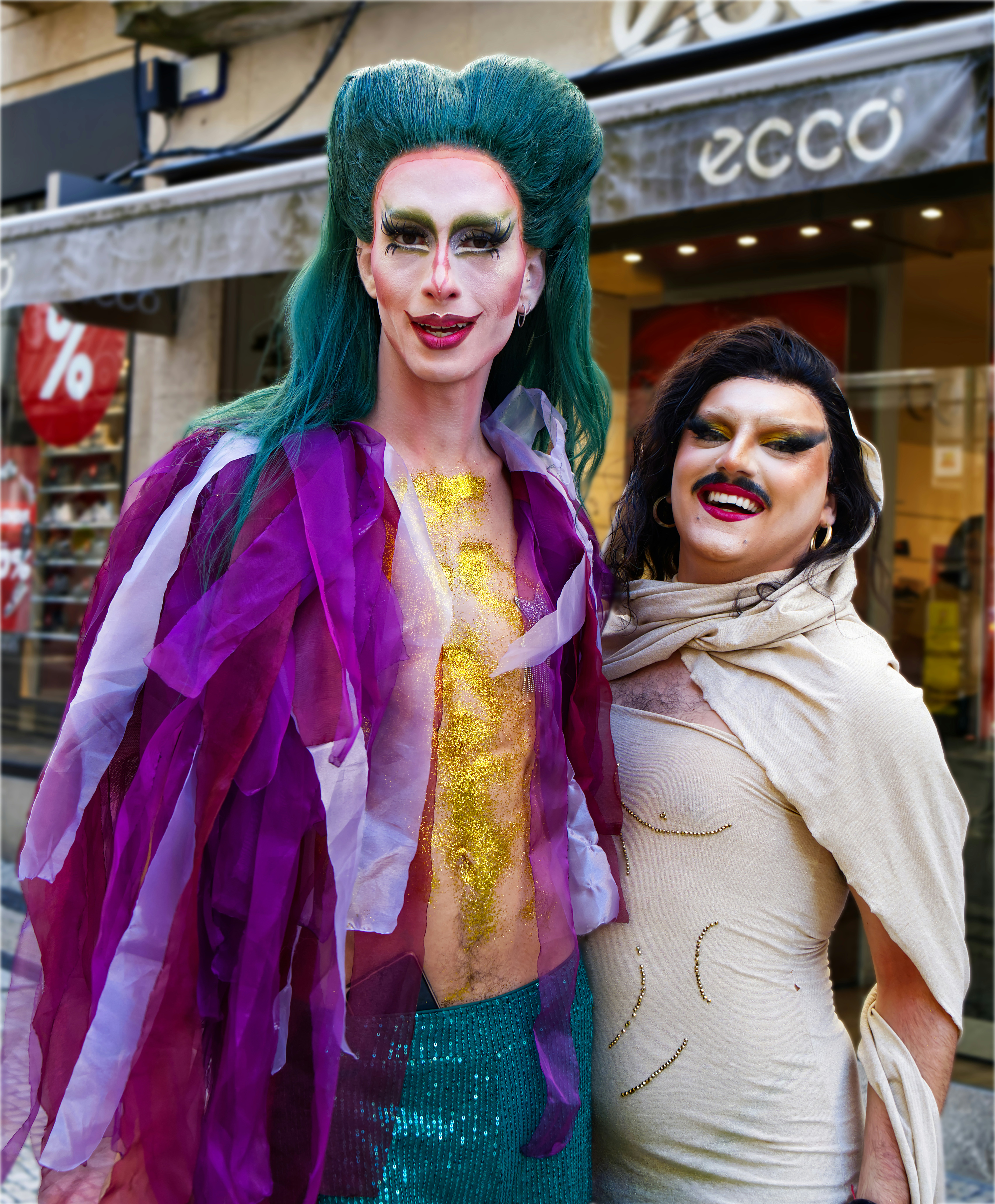 Rencontre flamboyante sous le ciel de Porto. Ce jour-là, la rue Sainte-Catherine, artère vibrante de la ville, s’est transformée en une scène éphémère où la couleur et l’audace ont pris le pas. Deux reines, drapées de lumière et de paillettes, ont croisé mon objectif. À gauche, une déesse aux cheveux verts émeraude, coiffés en une couronne de boucles théâtrales, enveloppée dans une cape violette et or qui semble capturer les reflets du soleil portugais. Son regard malicieux et ses lèvres pulpeuses, soulignées d’un rouge profond, racontent une histoire de fierté et de liberté. À ses côtés, une silhouette plus discrète mais tout aussi captivante, vêtue d’une robe beige ornée de motifs dorés, porte avec élégance une écharpe qui danse au rythme de la brise urbaine.