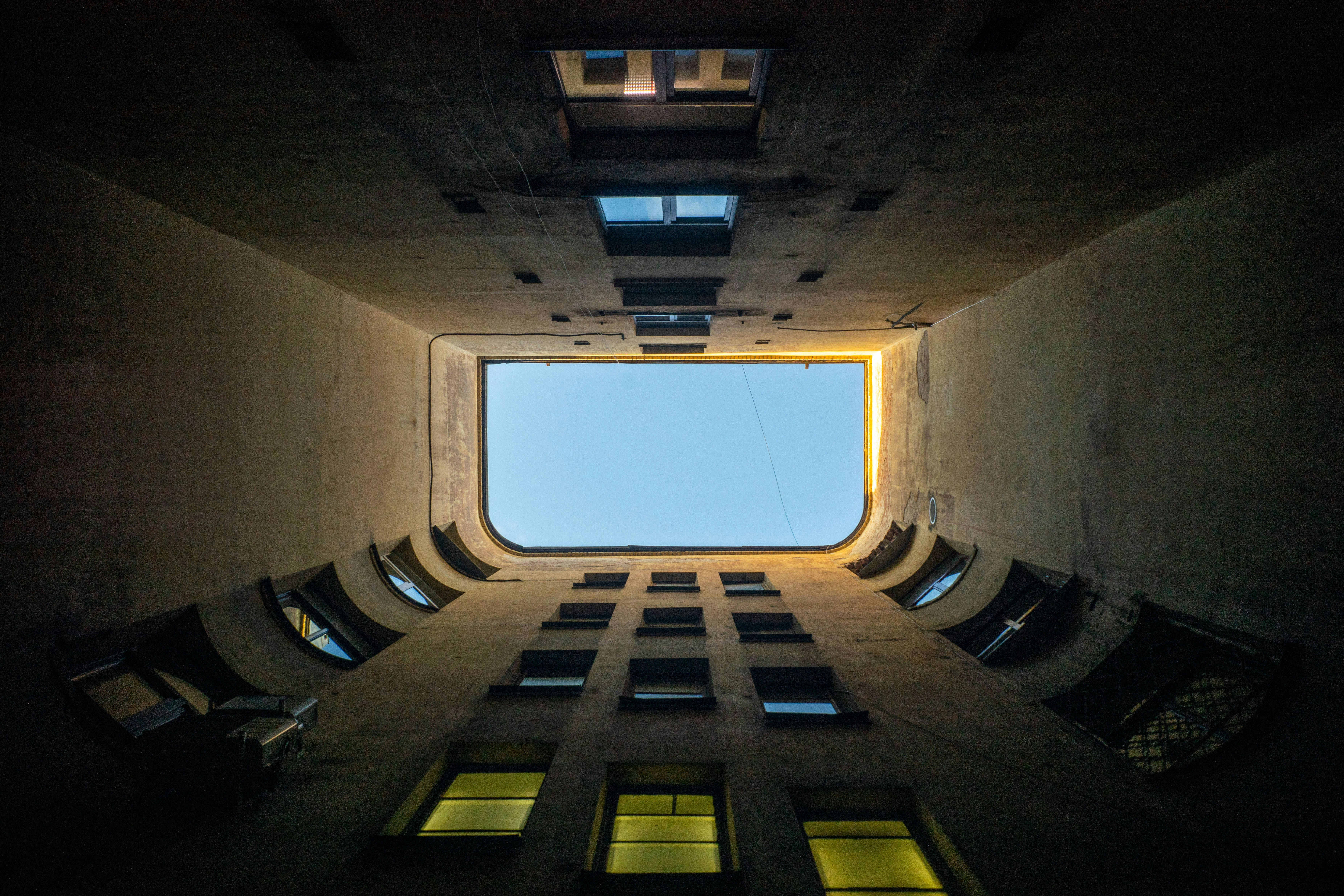 Converging walls frame a rectangular opening toward a pale blue sky; the shot emphasizes vertical urban geometry and the glow of internal window lights.