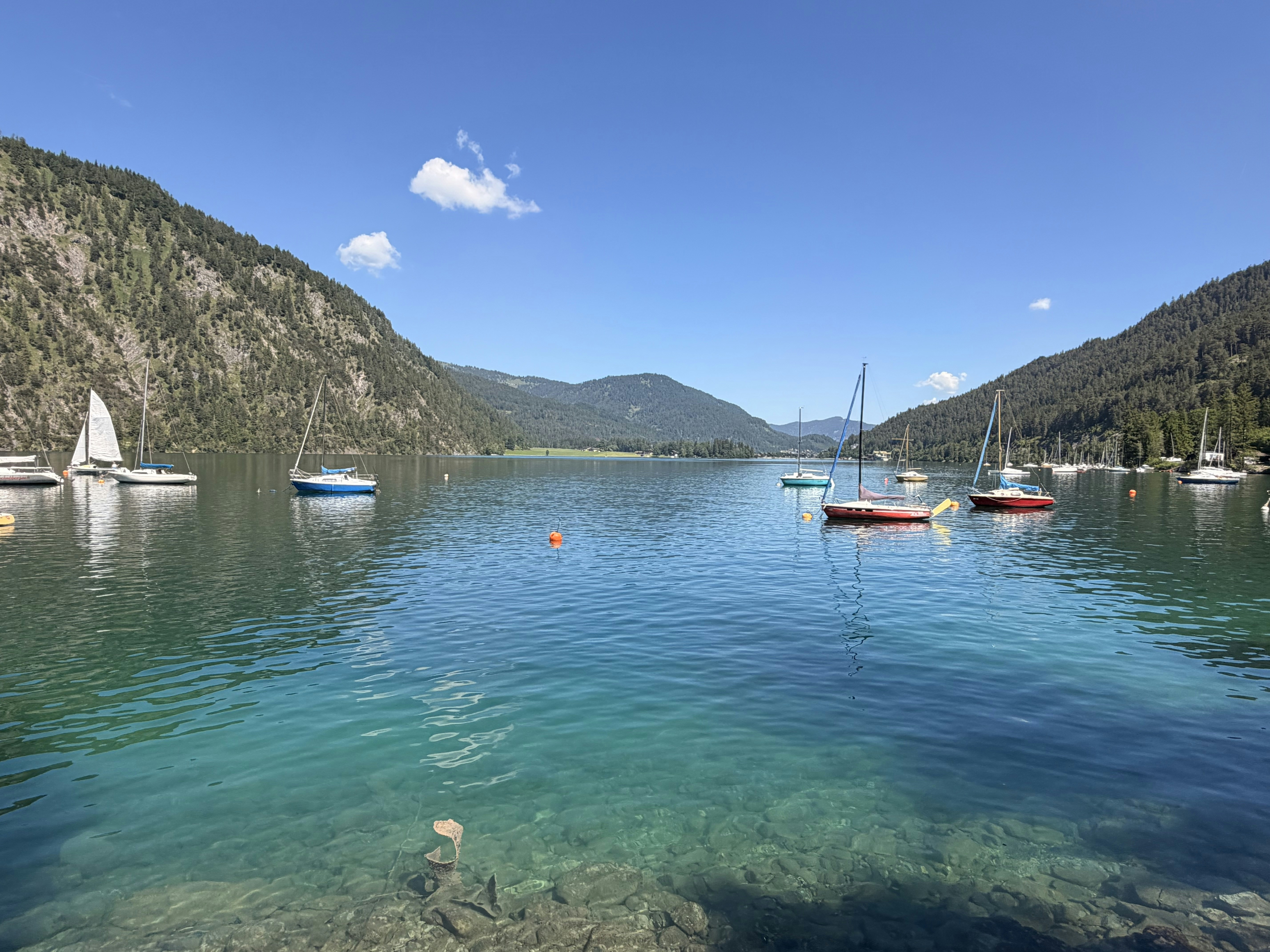 Clear alpine lake with anchored sailboats and turquoise shallow waters near the shore, framed by forested mountains under a bright blue sky.