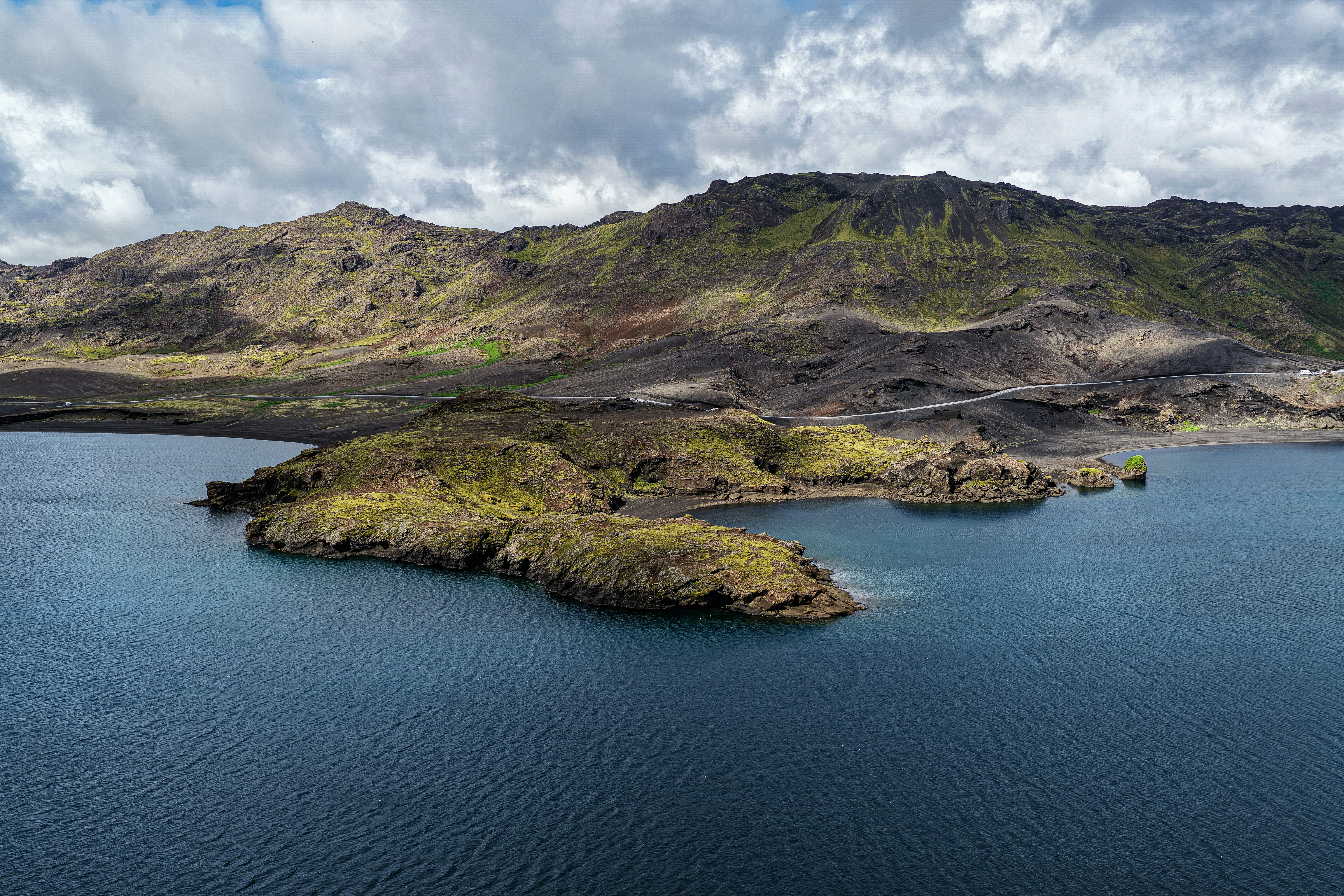 Coastal landscape with a rugged island connected by a winding causeway, surrounded by deep blue water and mossy hills under a partly cloudy sky.