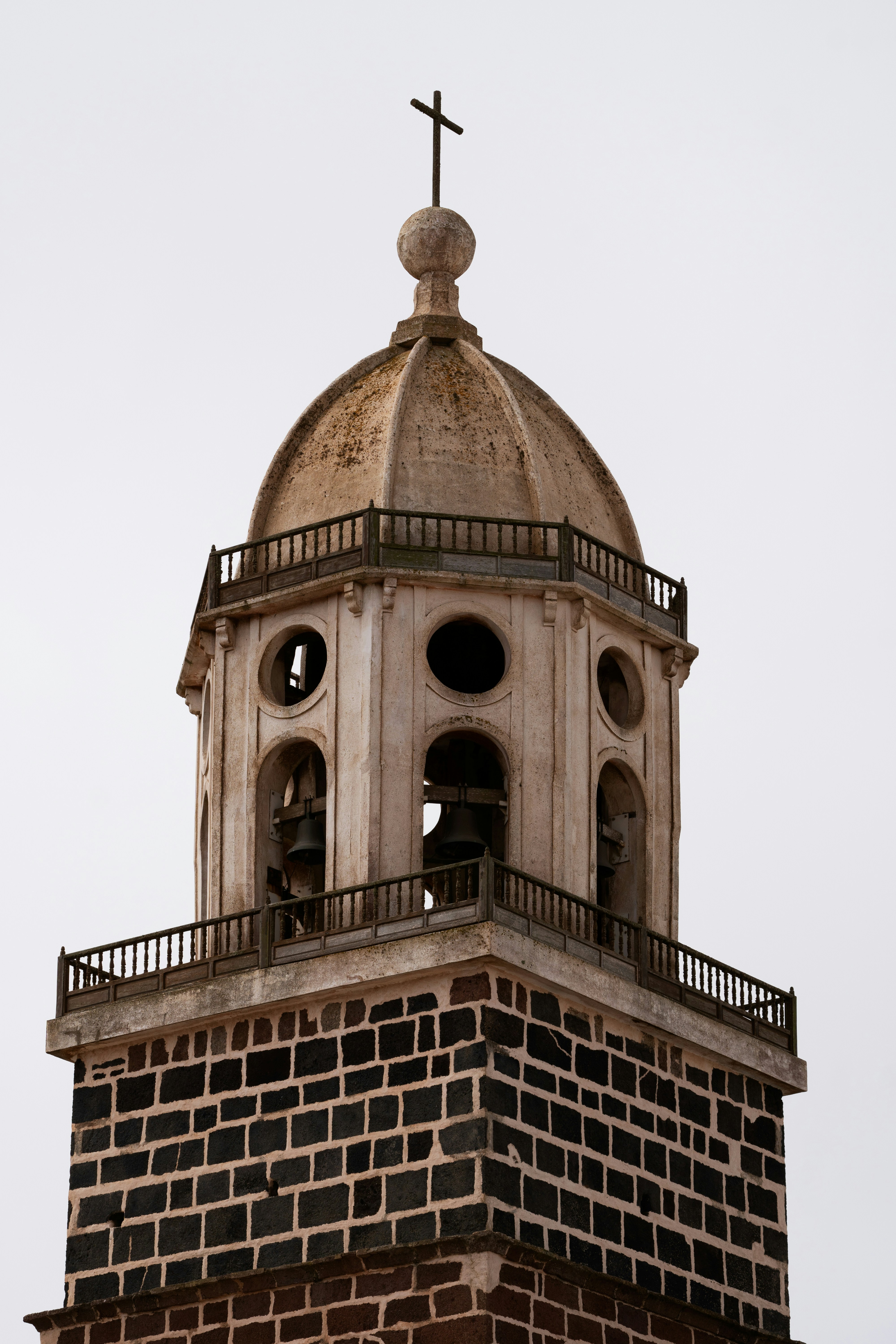 A historic stone bell tower rises against a pale sky, featuring arched openings and a brick base with wrought-iron railings. The structure stands tall and solitary, emphasizing architectural detail.