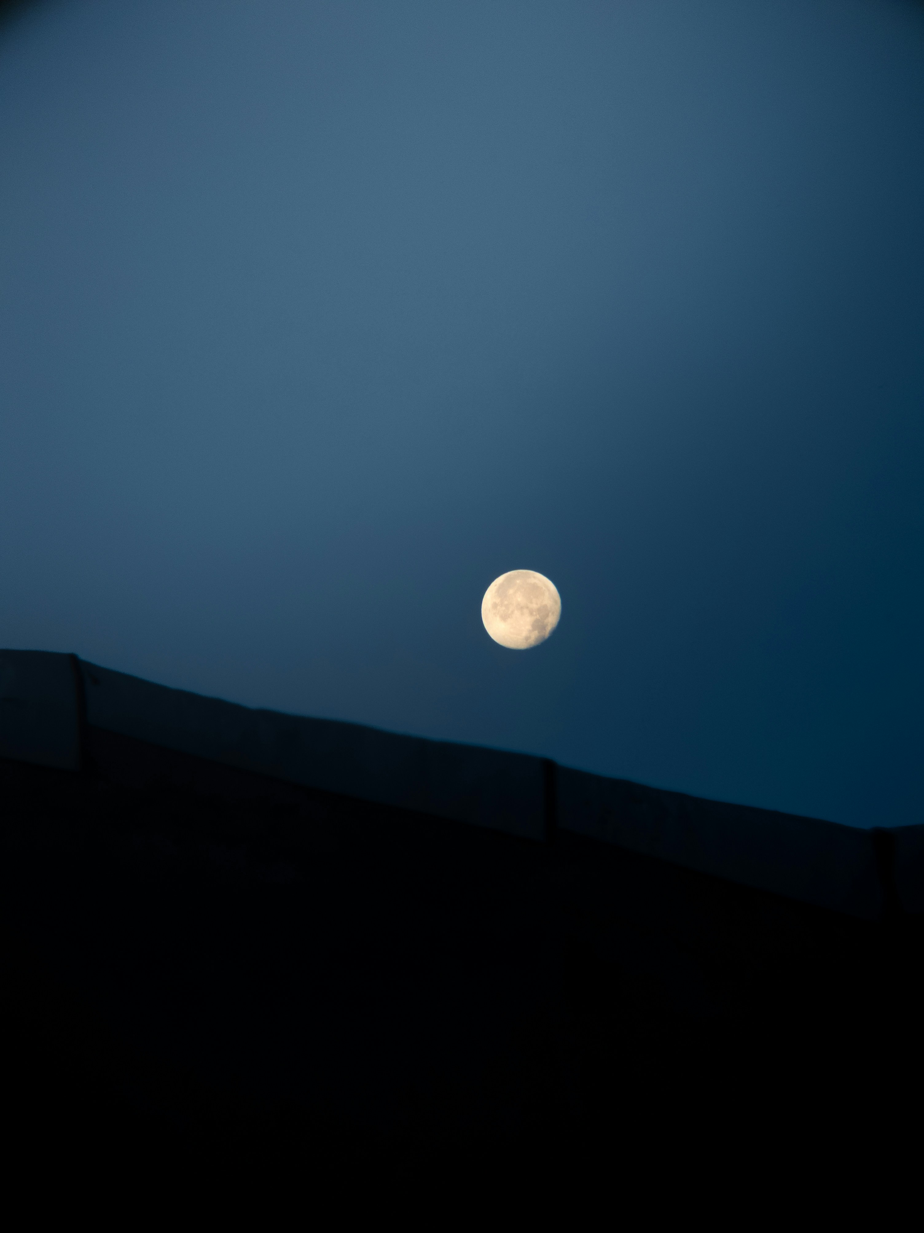 Moon glows softly above a dark rooftop silhouette against a deep blue twilight sky. A minimal urban scene with emphasis on the luminous lunar disk.