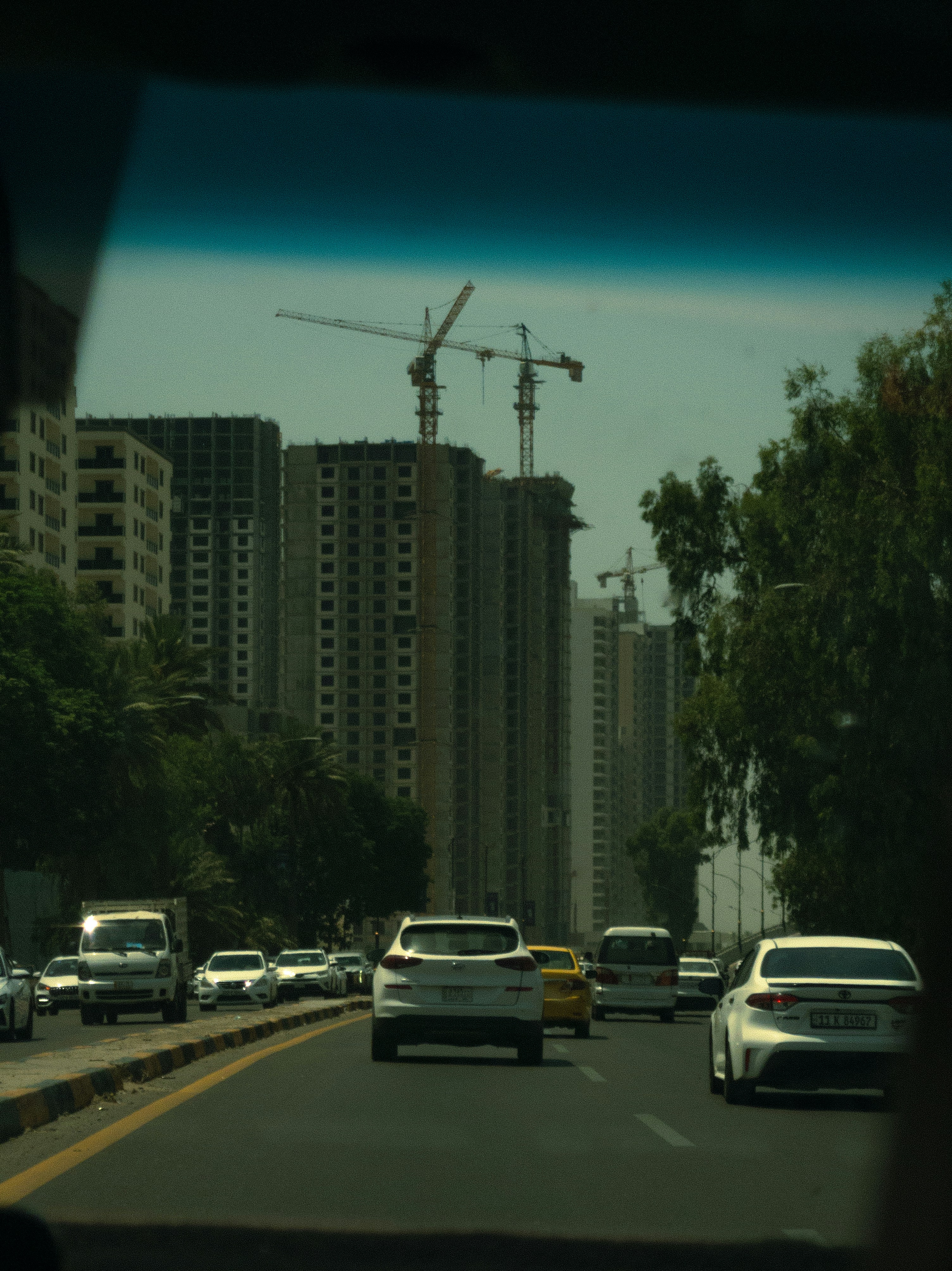Busy city street with vehicles in motion beneath a cluster of high-rise construction cranes against a tree-lined backdrop.