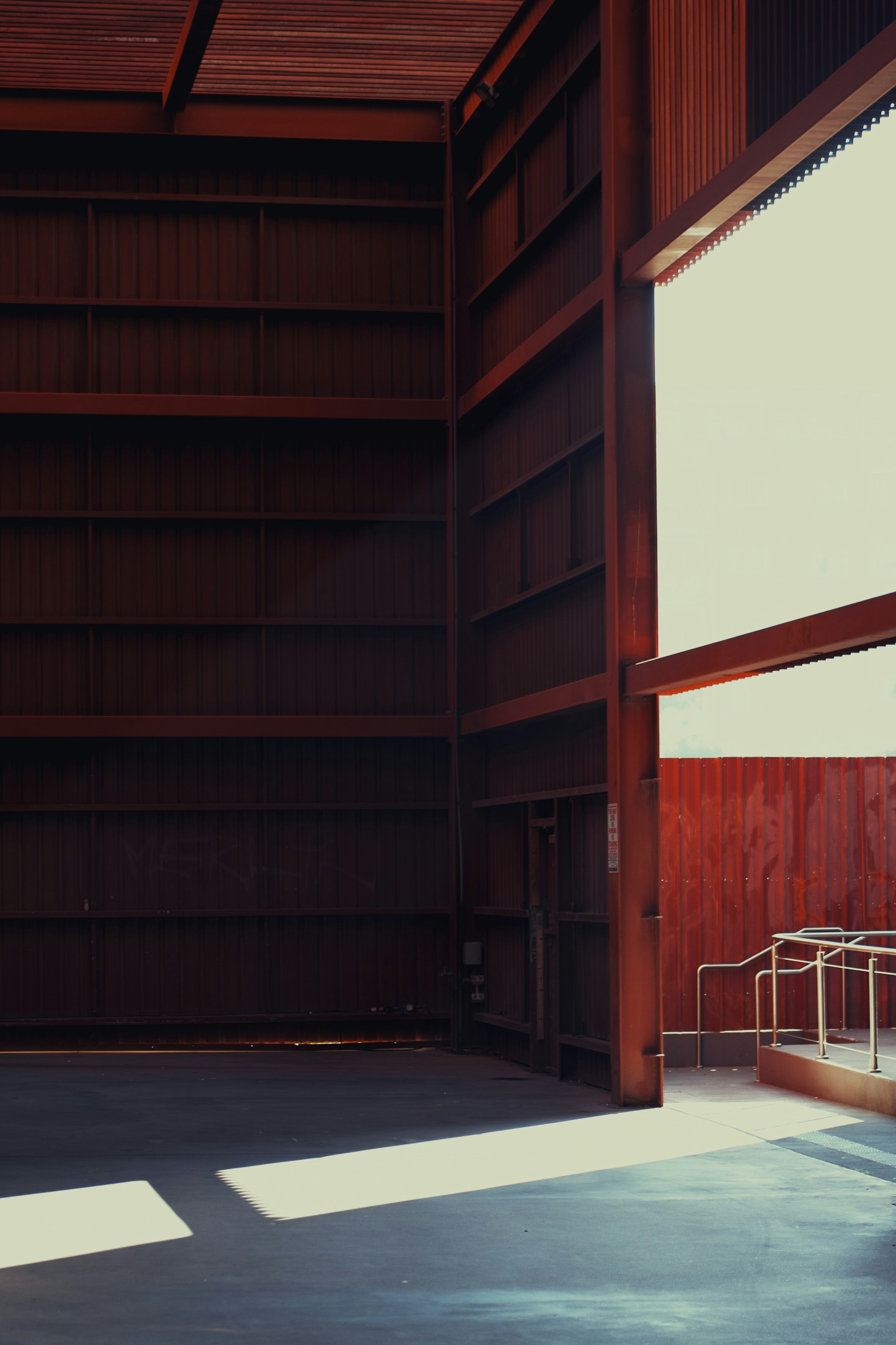 Sunlit interior of a large, open warehouse with dark red-orange framework; strong light pours in from the right, casting geometric shadows across the concrete floor and highlighting the stark contrast between interior structure and exterior fence.