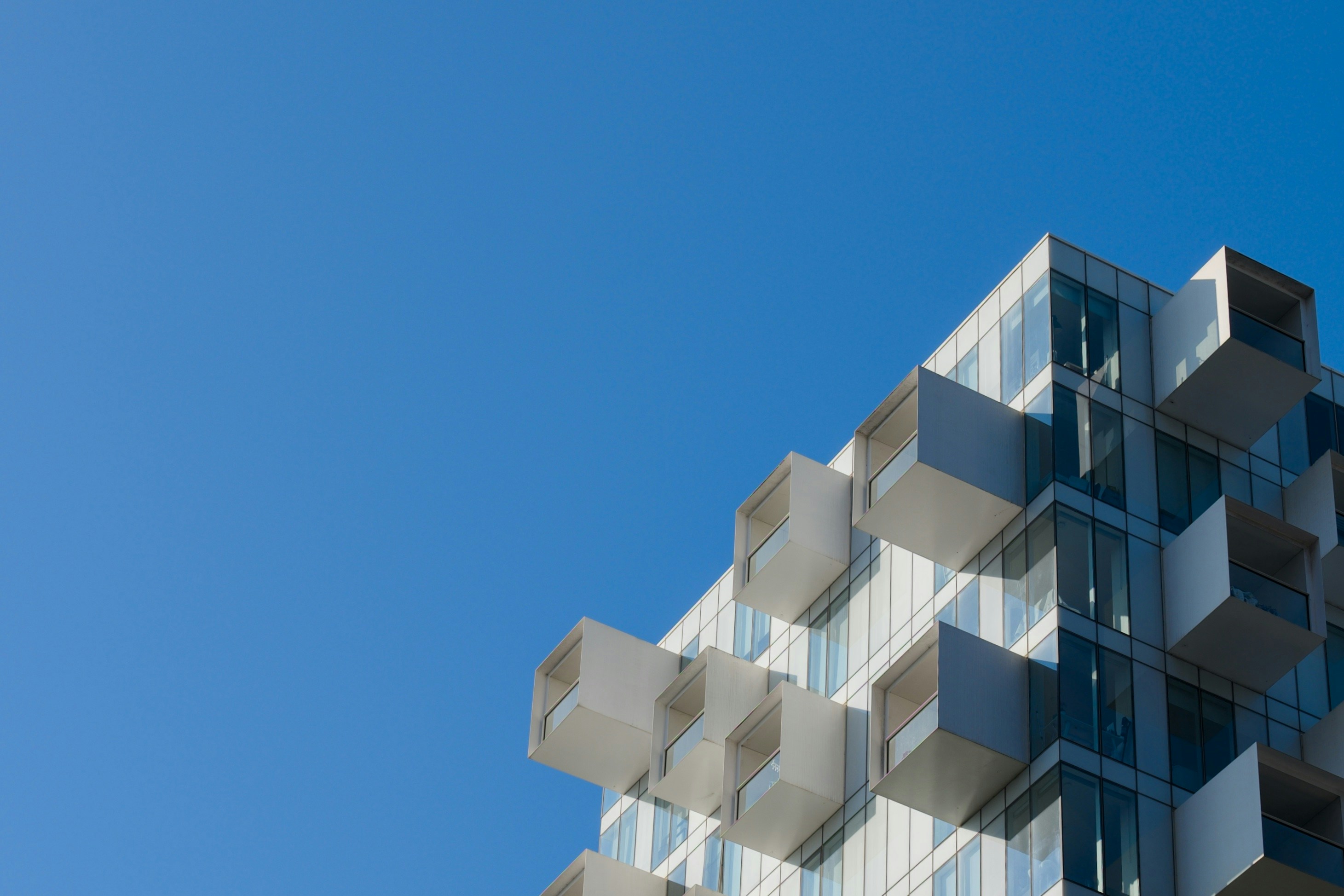 Sleek glass-and-concrete building corner with stacked balconies against a vivid blue sky; architectural geometry and light play emphasize depth and structure.