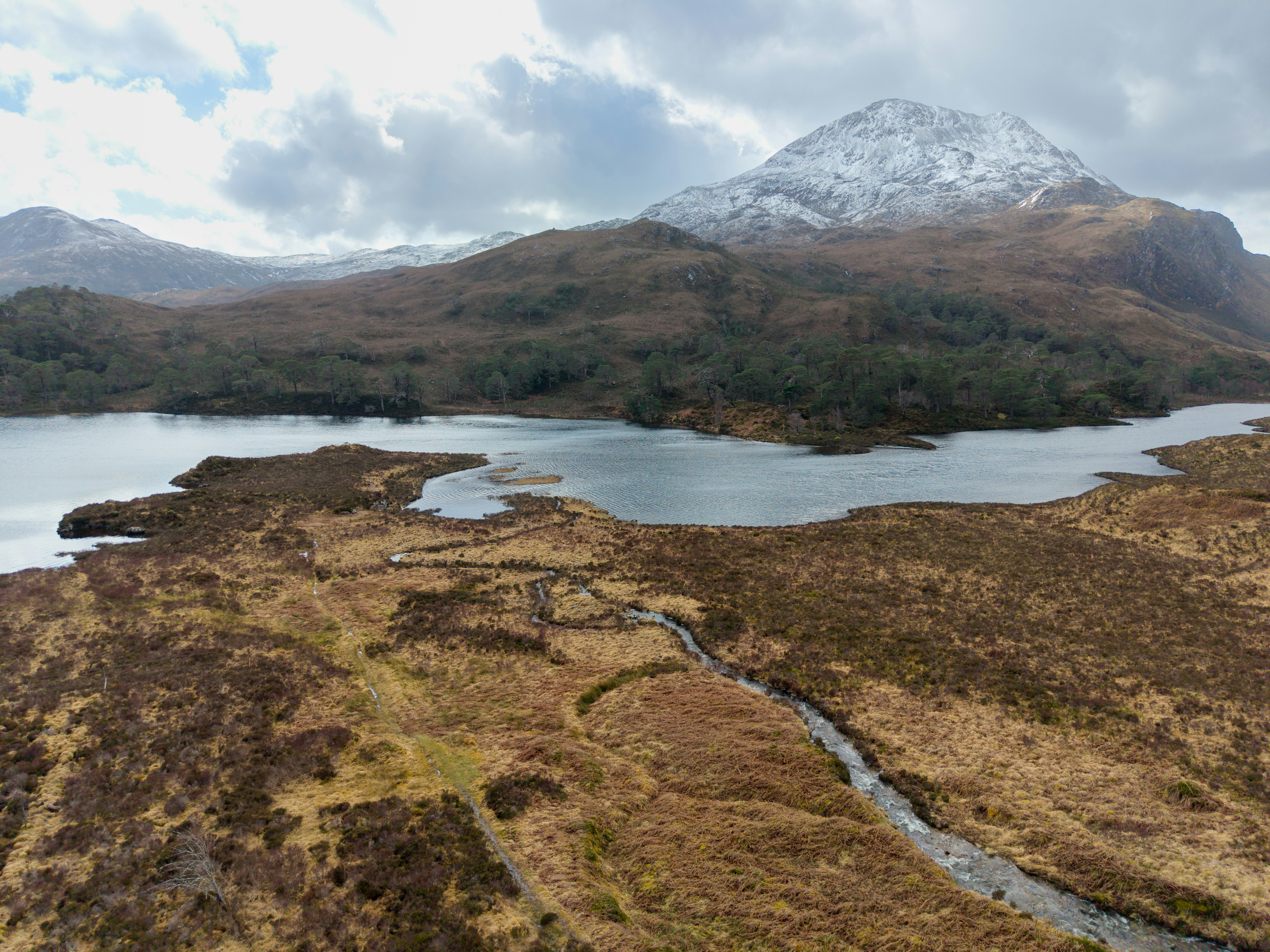 Moorland foreground with a winding stream leading to a distant snow-topped peak and a calm lake; natural landscape scene capturing wide terrain and distant ridge.