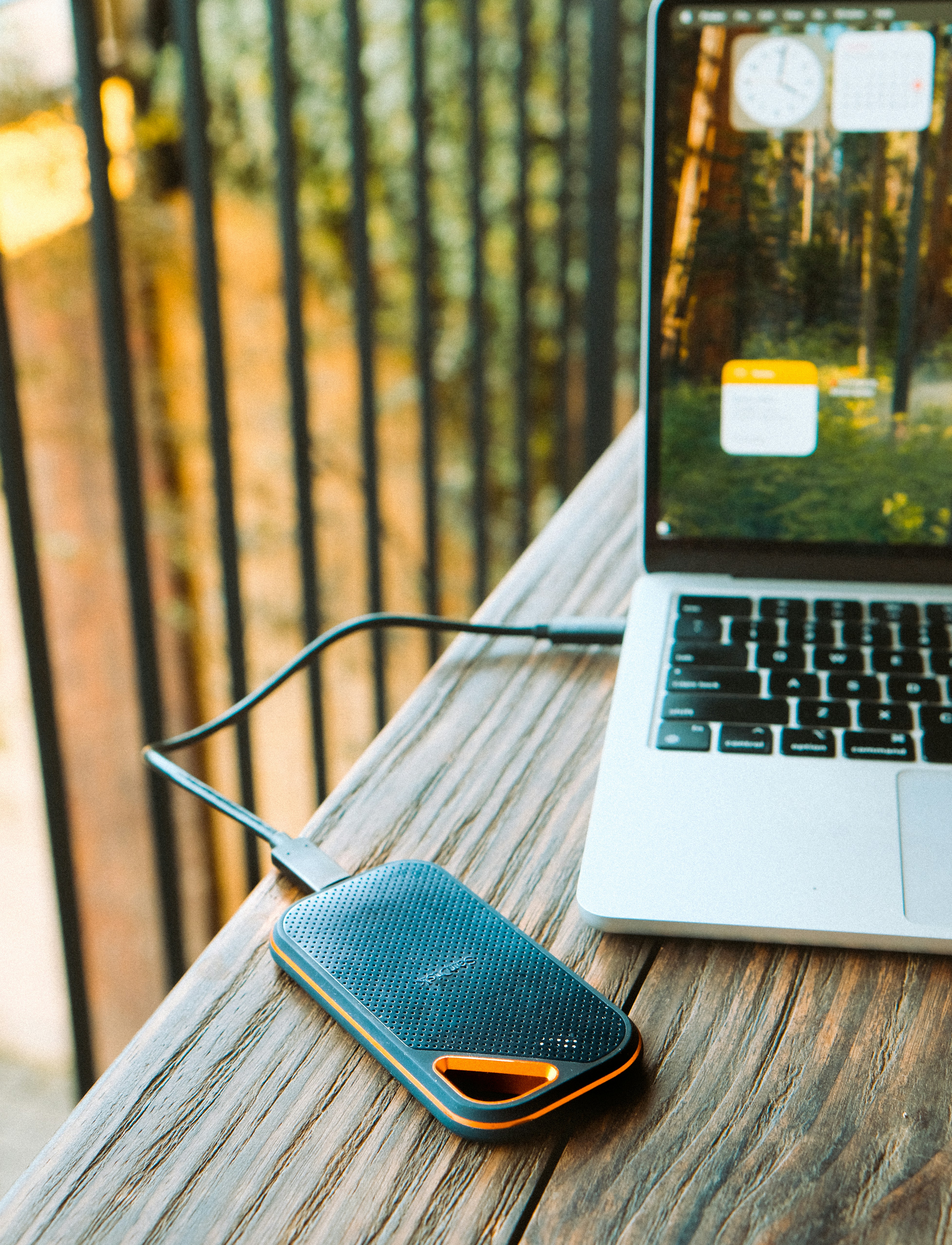 Compact external hard drive rests on a weathered wooden table beside a laptop displaying a nature scene, with warm sunset tones and vertical balcony bars in the background.