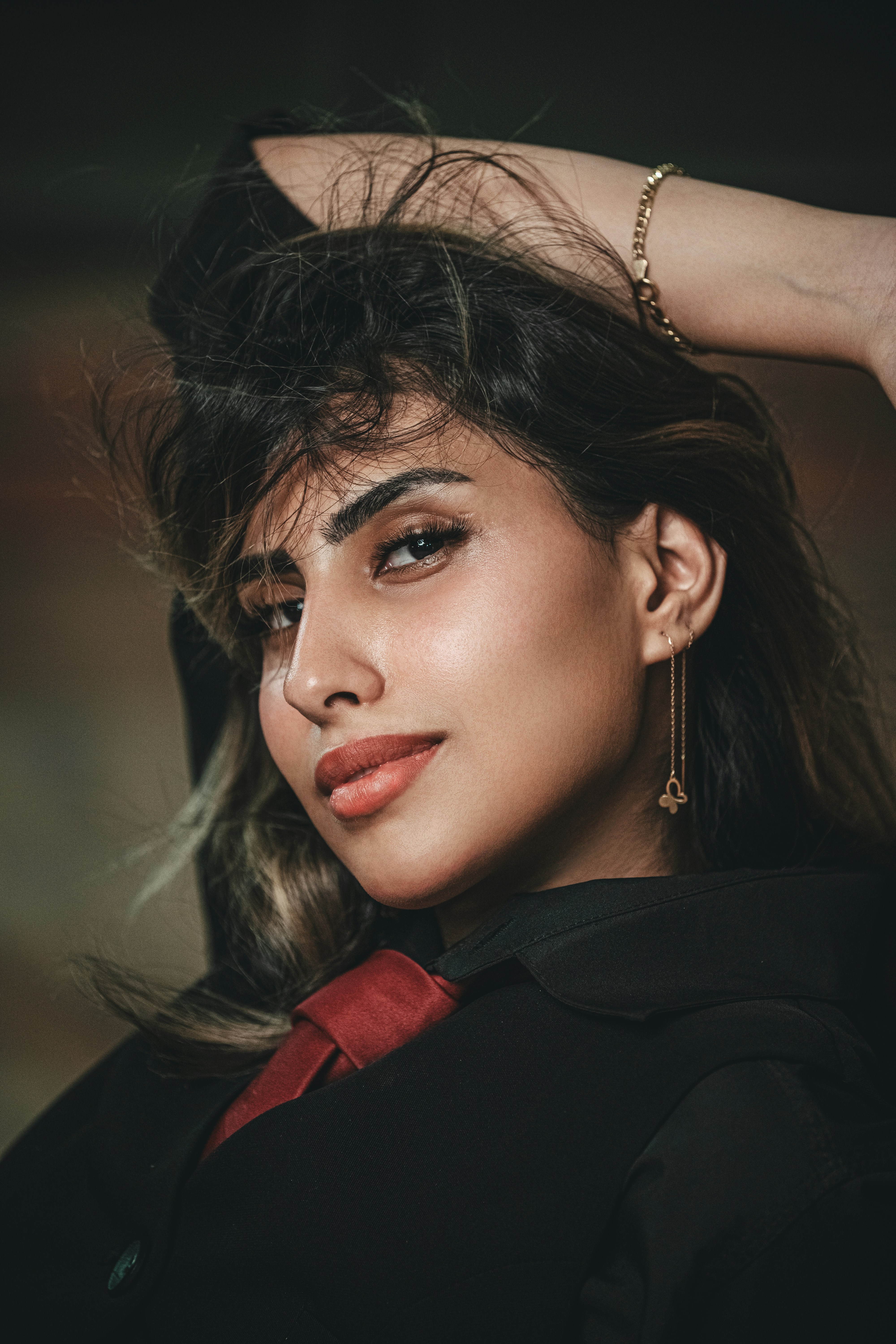 Close-up portrait of a woman with warm skin tones and subtle makeup, under controlled lighting; a glossy black outfit and red tie add contrast. This is a stylized studio photograph.