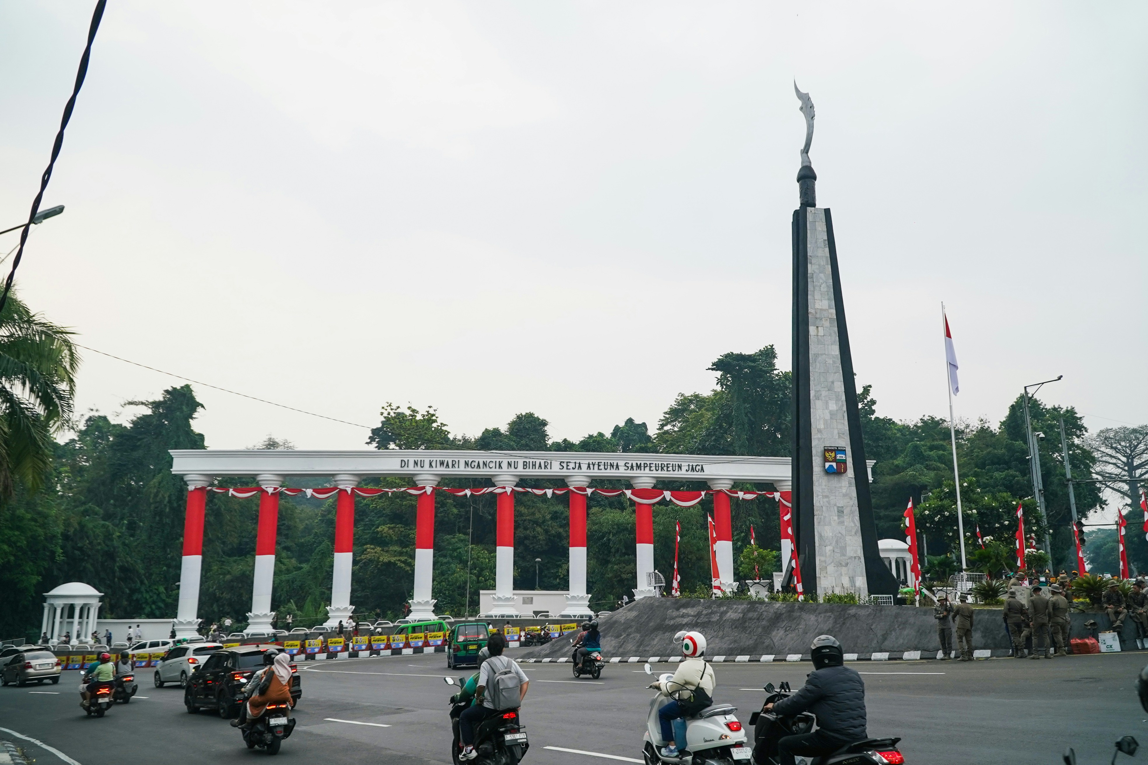 A busy roundabout with bold red-and-white colonnades surrounding a central monument and a tall obelisk; motorbikes traverse the foreground under an overcast sky. The scene captures urban ceremony and traffic convergence around a civic landmark.