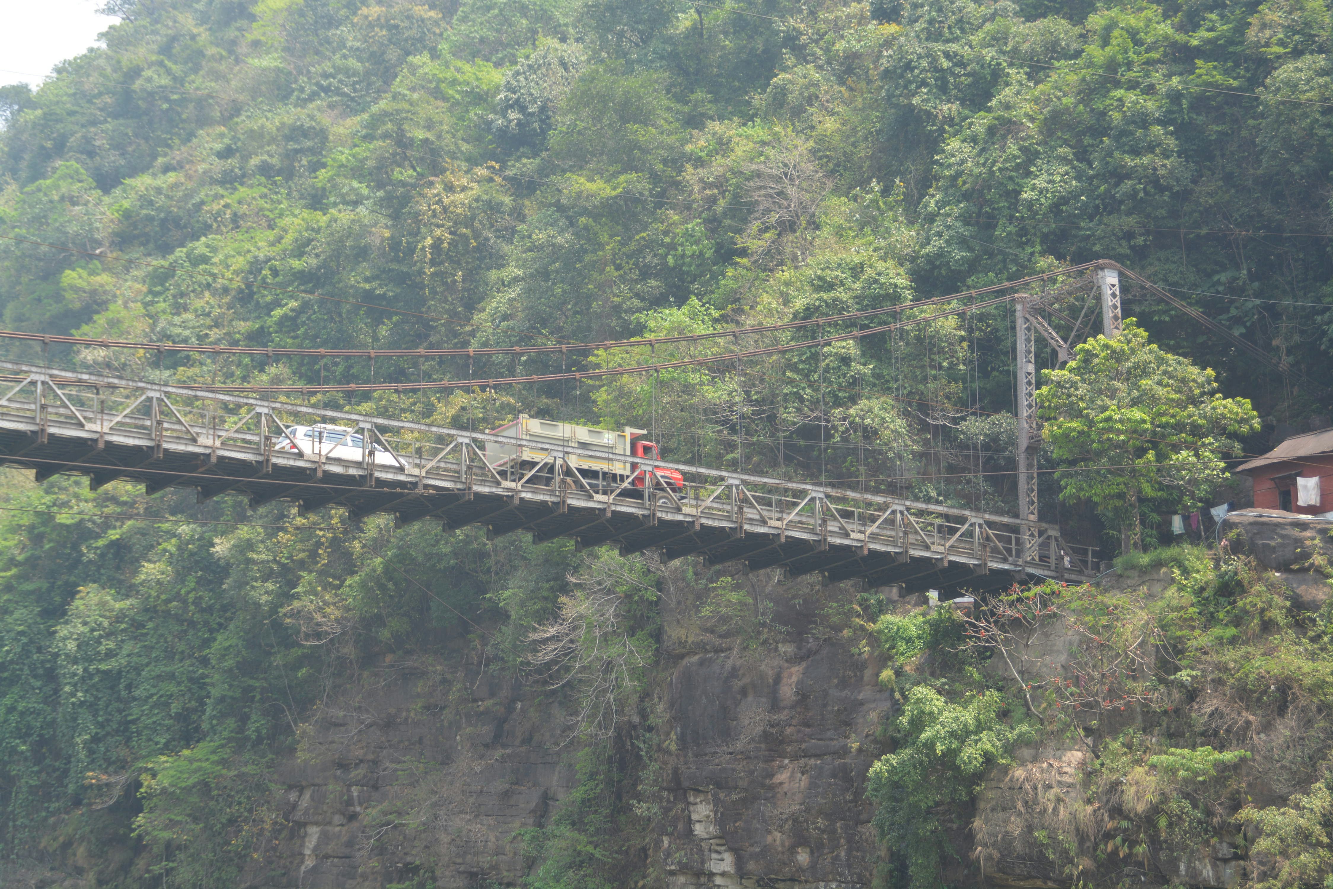 Bridge over a forested gorge with a vehicle and crane traversing the span, captured from the hillside. Aerial rail lines and pylons hint at a suspended crossing.