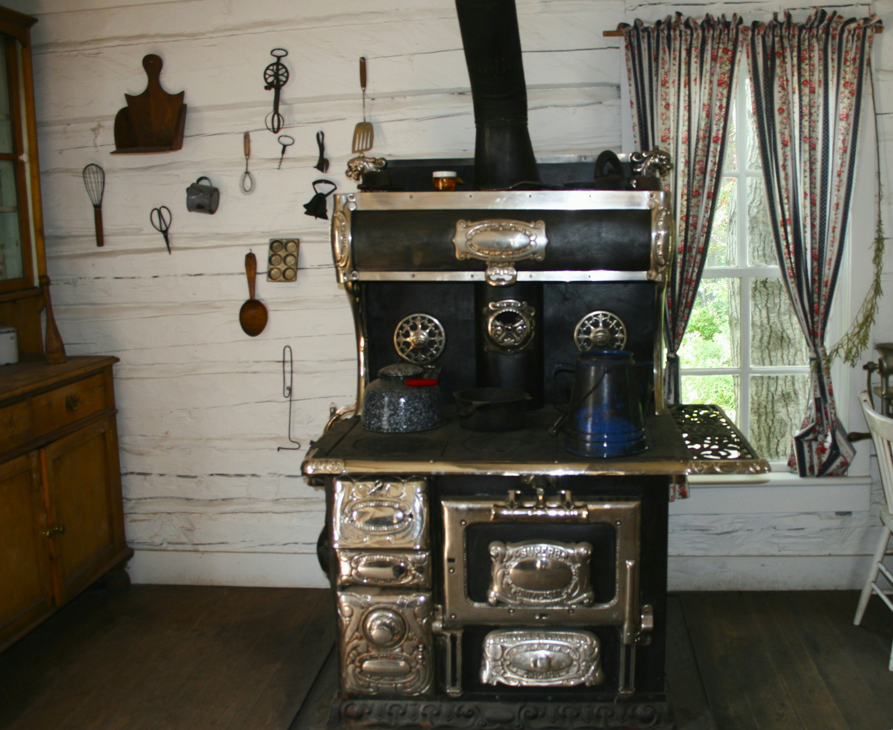 A beautifully preserved, antique black cast iron cook stove serves as the focal point of a rustic, historic log cabin kitchen at Fort Edmonton Park in Alberta, Canada. The ornate vintage stove features polished chrome details, a raised warming shelf, an old speckled kettle, and a classic blue enamel coffee pot resting on the stovetop. Against the white-washed log walls, a variety of pioneer culinary tools and iron utensils hang neatly on display, including a wooden paddle, whisks, and cooking spoons. To the right, a multi-pane glass window dressed in patterned floral curtains allows soft dayli