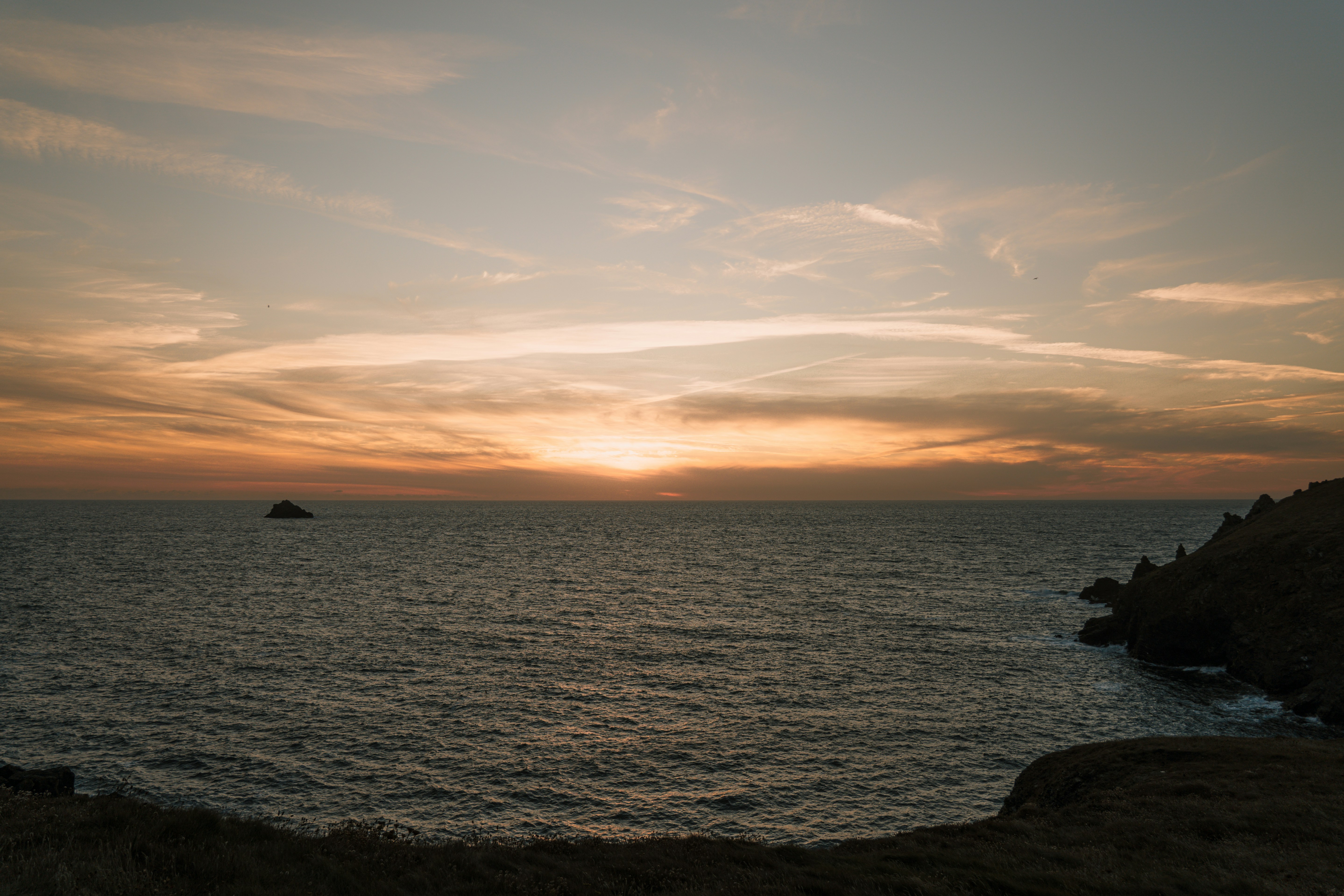 Soft sunset over a quiet sea with a rocky shoreline, the sun dipping near the horizon and a small rock silhouette breaking the calm water.