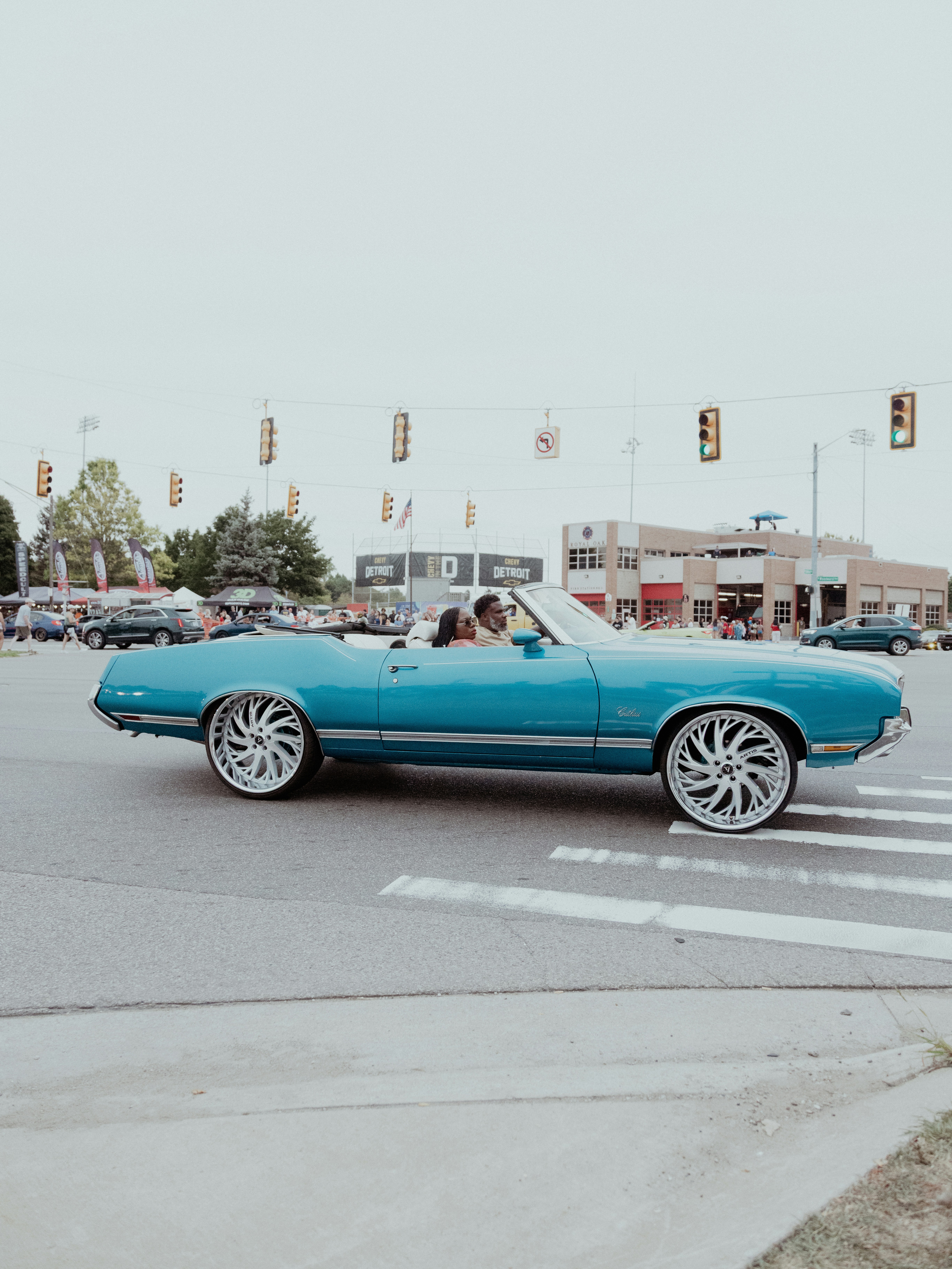 A classic Chevrolet Chevelle convertible cruising through Detroit, finished in vibrant turquoise with oversized chrome wheels. Captured at an intersection with the city, crowds, and summer car culture in the background. A blend of classic American design and modern custom style. Tags: Chevrolet, Chevelle, classic car, convertible, Detroit, Michigan, car culture, turquoise, vintage car, American muscle, automotive, summer, street photography, chrome wheels, custom car, cityscape