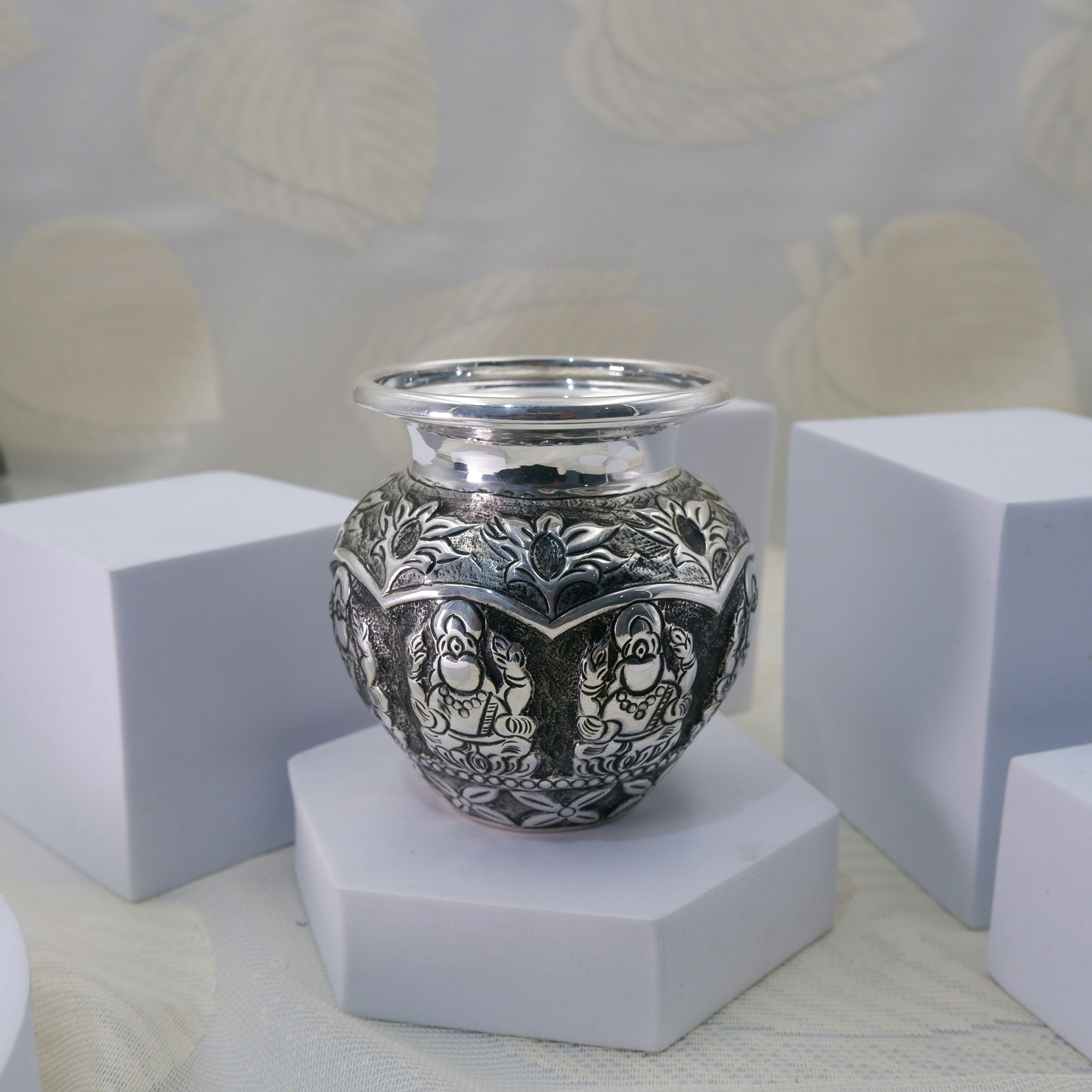 Intricately engraved silver vessel sits atop a pale pedestal, flanked by pale geometric blocks in a softly lit setting. A decorative object-focused shot highlighting craftsmanship.