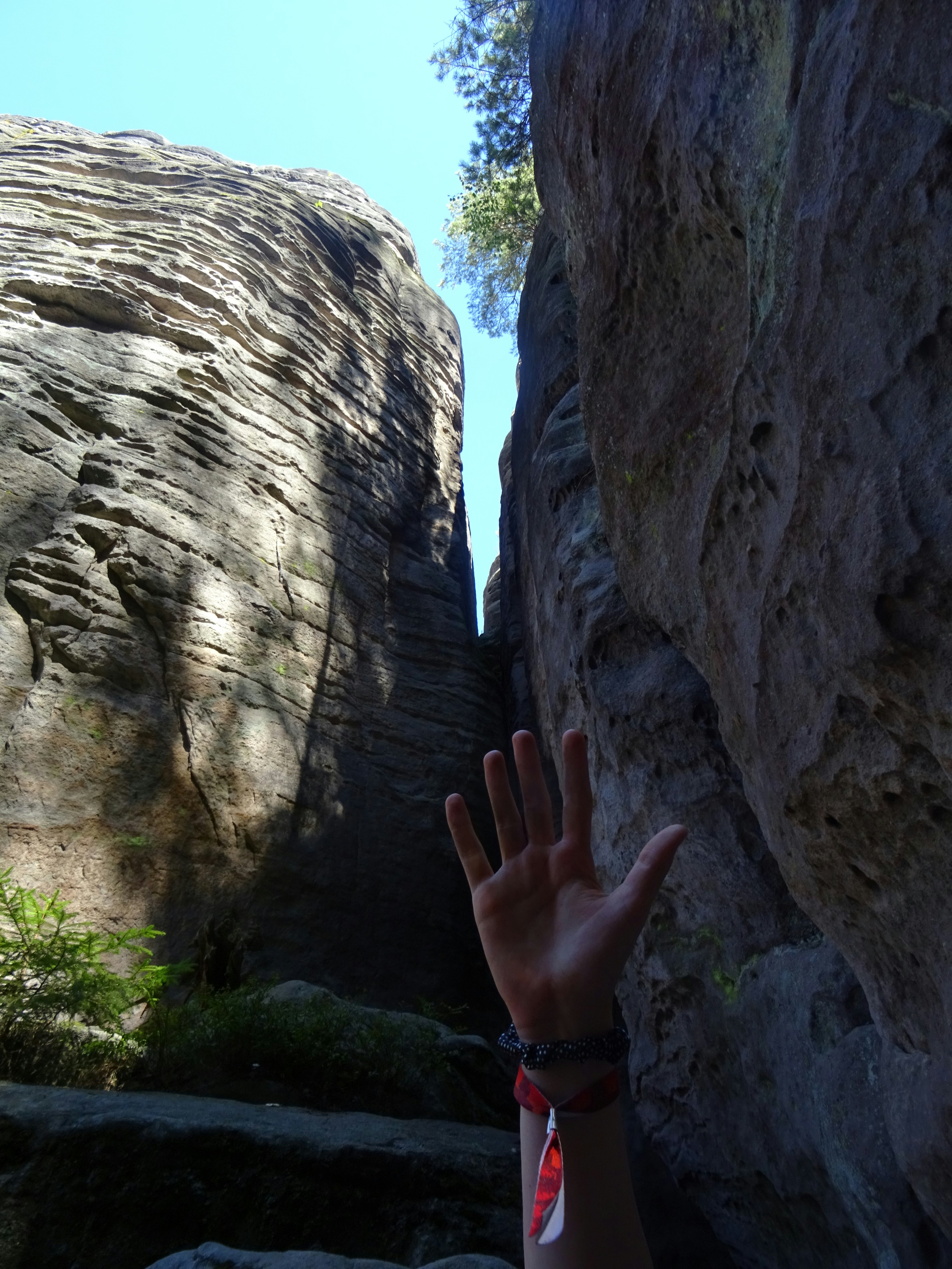 A lone arm and hand extend upward from the bottom edge toward a sky-lit gap between towering rock formations; the scene emphasizes scale and the contrast between shadowed walls and bright sky. This is a natural landscape with a captured human element.