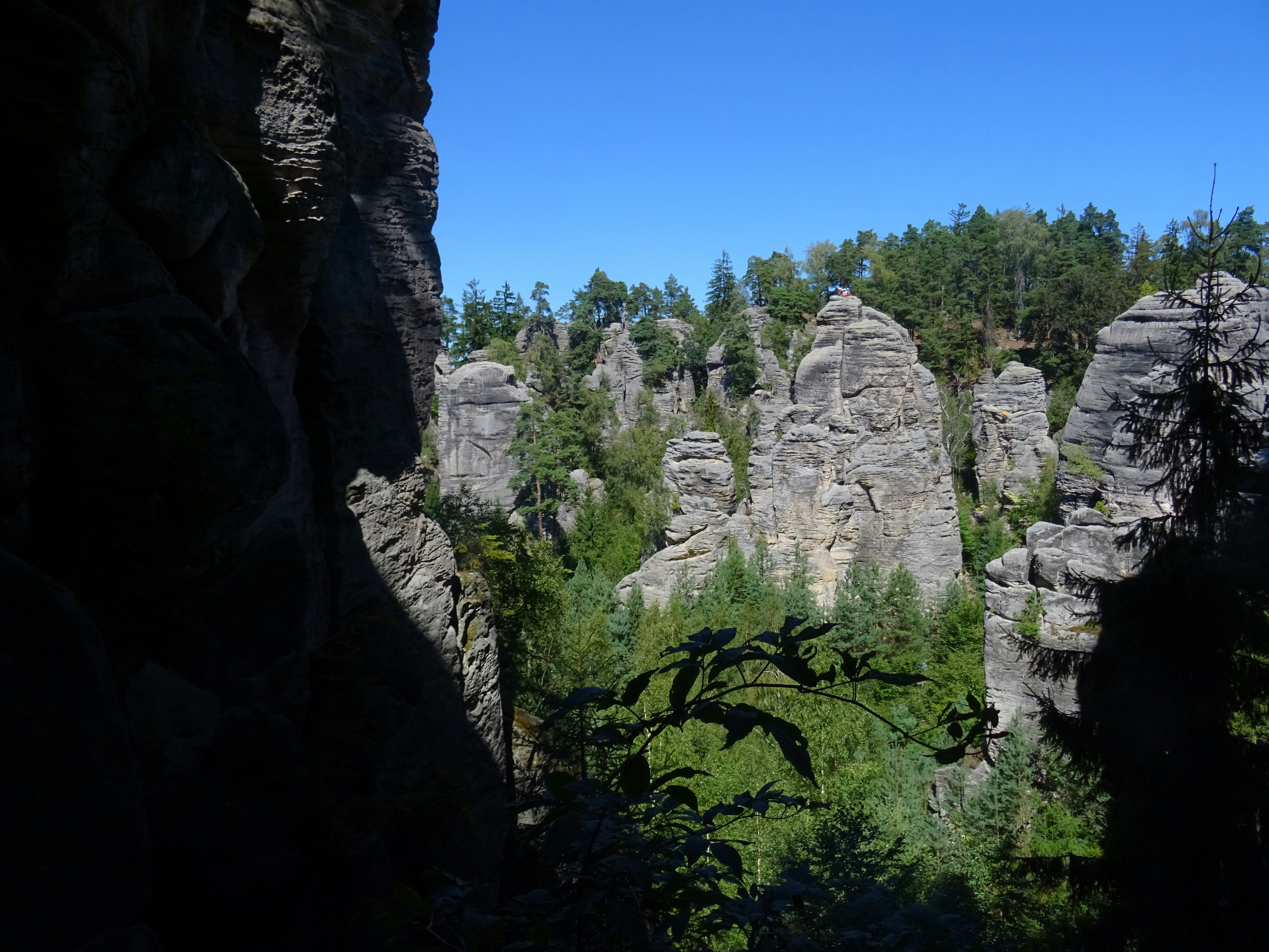 Dramatic rock pillars rise above a green forest under a bright blue sky, with a dark foreground cliff creating a natural frame.