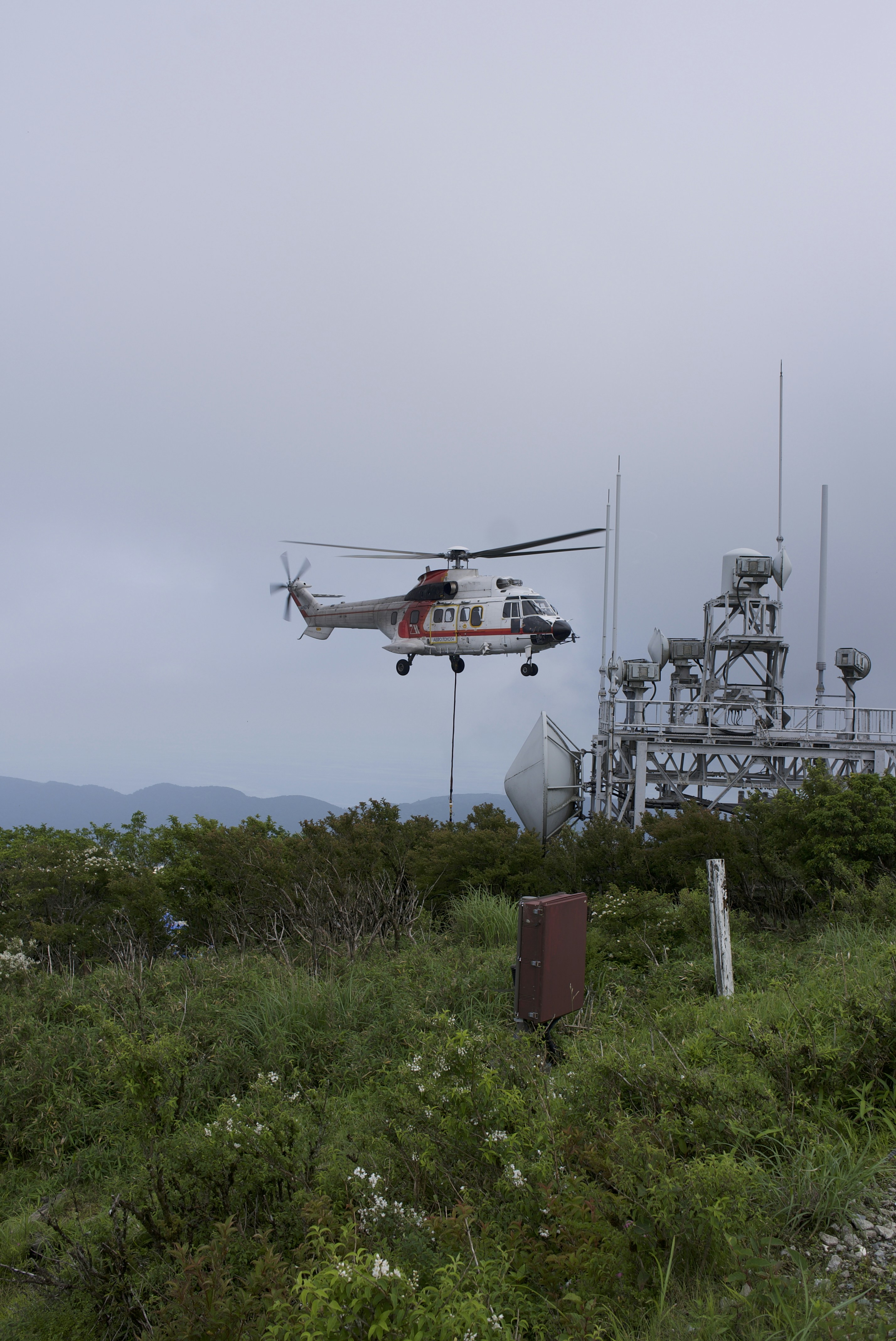 Color-saturated helicopter hovers near a tall communications tower above a scrubby coastal landscape, with distant mountains on the horizon.