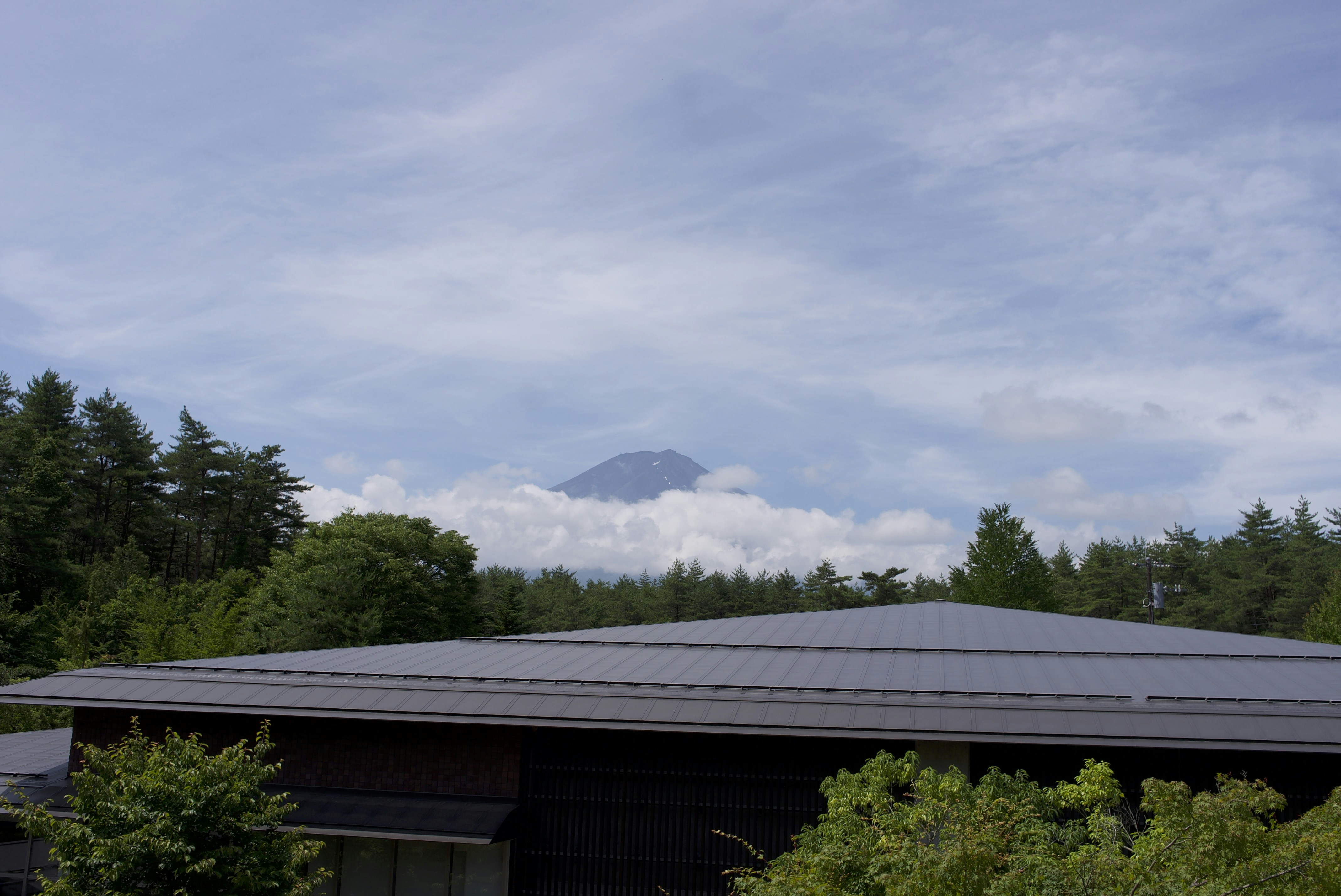 Low-angle view of rooftops with a distant mountain peak emerging from clouds, framed by trees and a bright sky. A calm landscape scene with a modern building foreground.