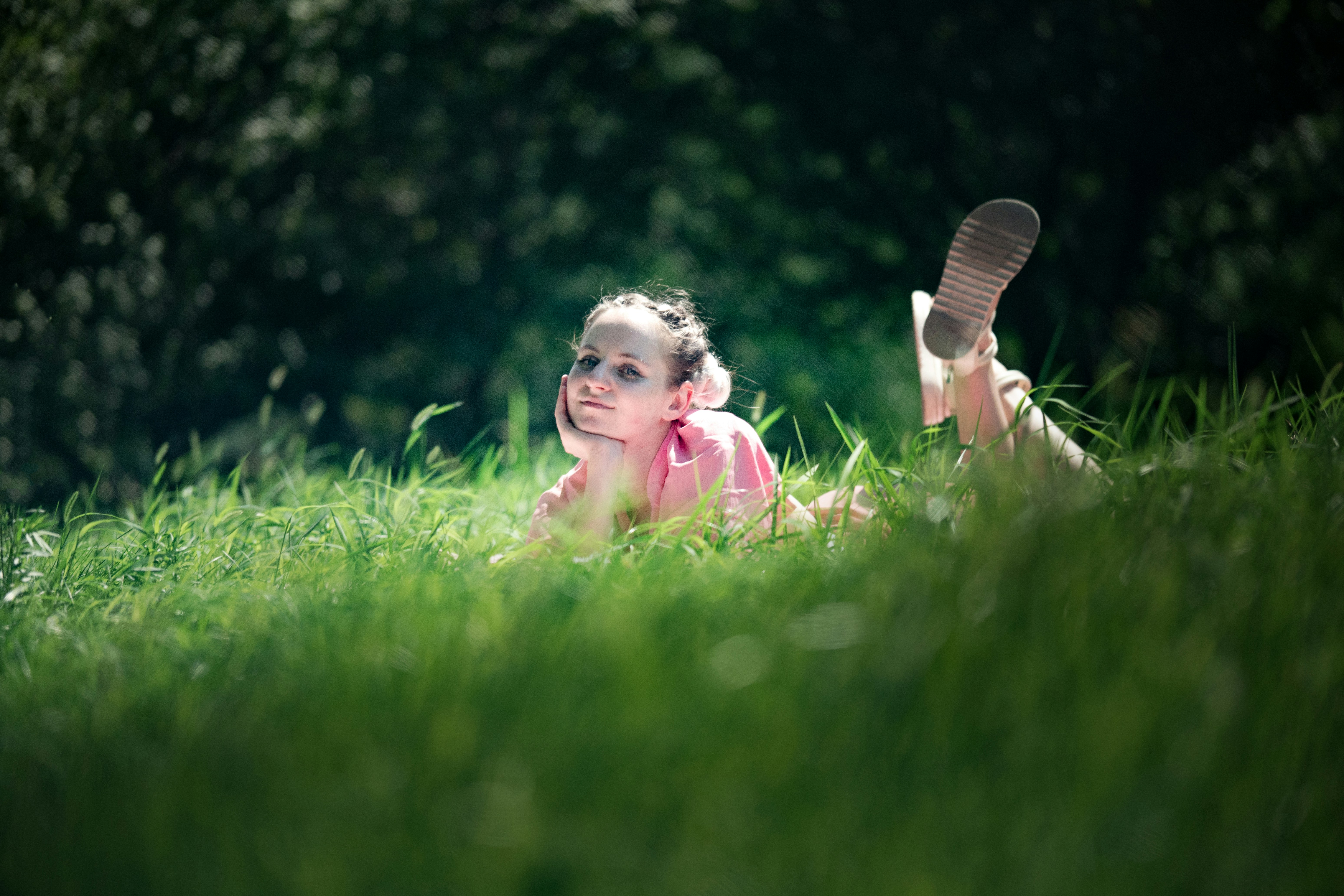 Child lying on grass with face and pink shirt visible, one leg kicked up; soft bokeh and natural light suggesting outdoor leisure and a candid moment.