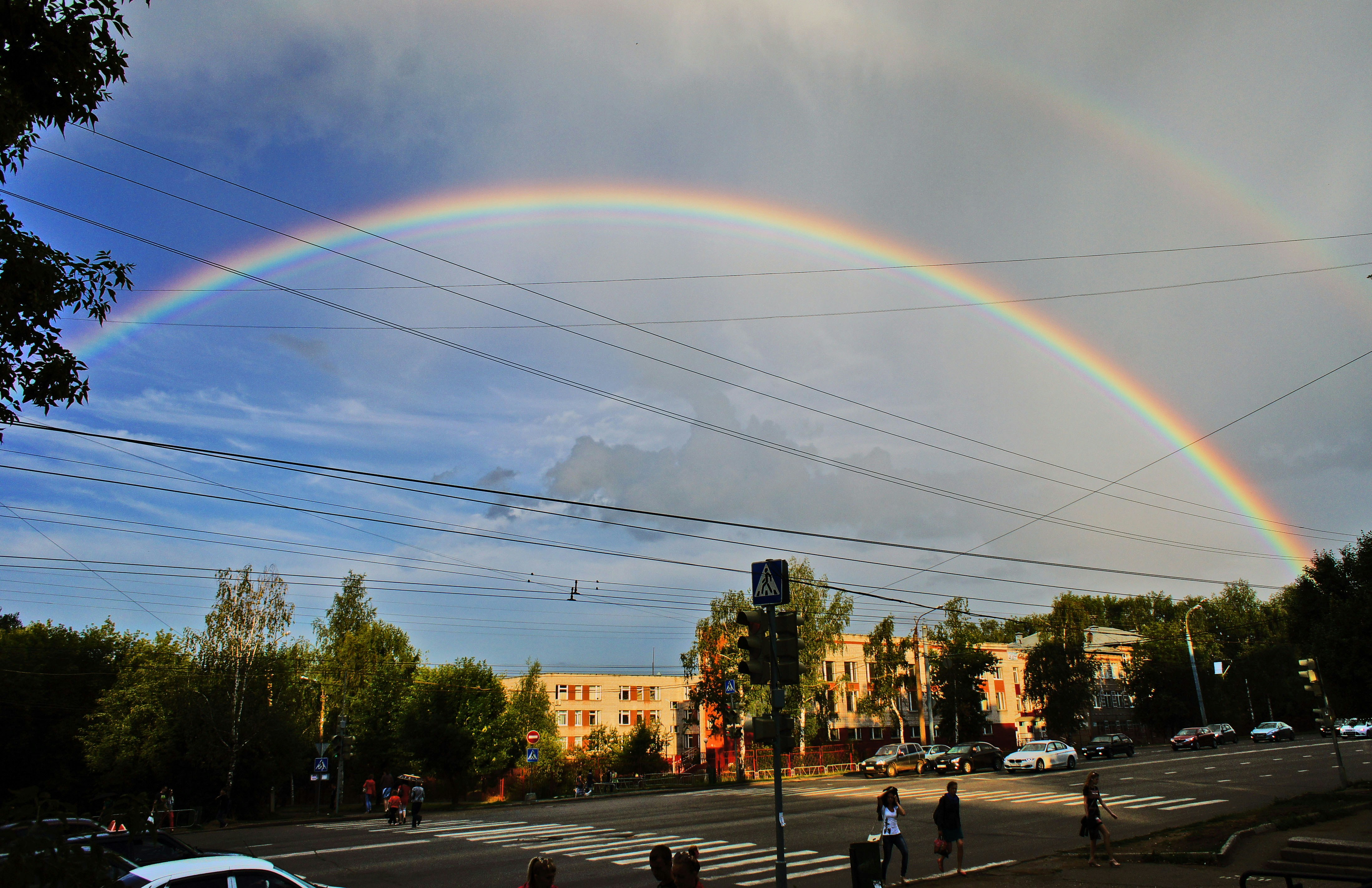 Vibrant double rainbow arches over a busy urban intersection with pedestrians and cars, captured at twilight with a clear sky and scattered clouds.