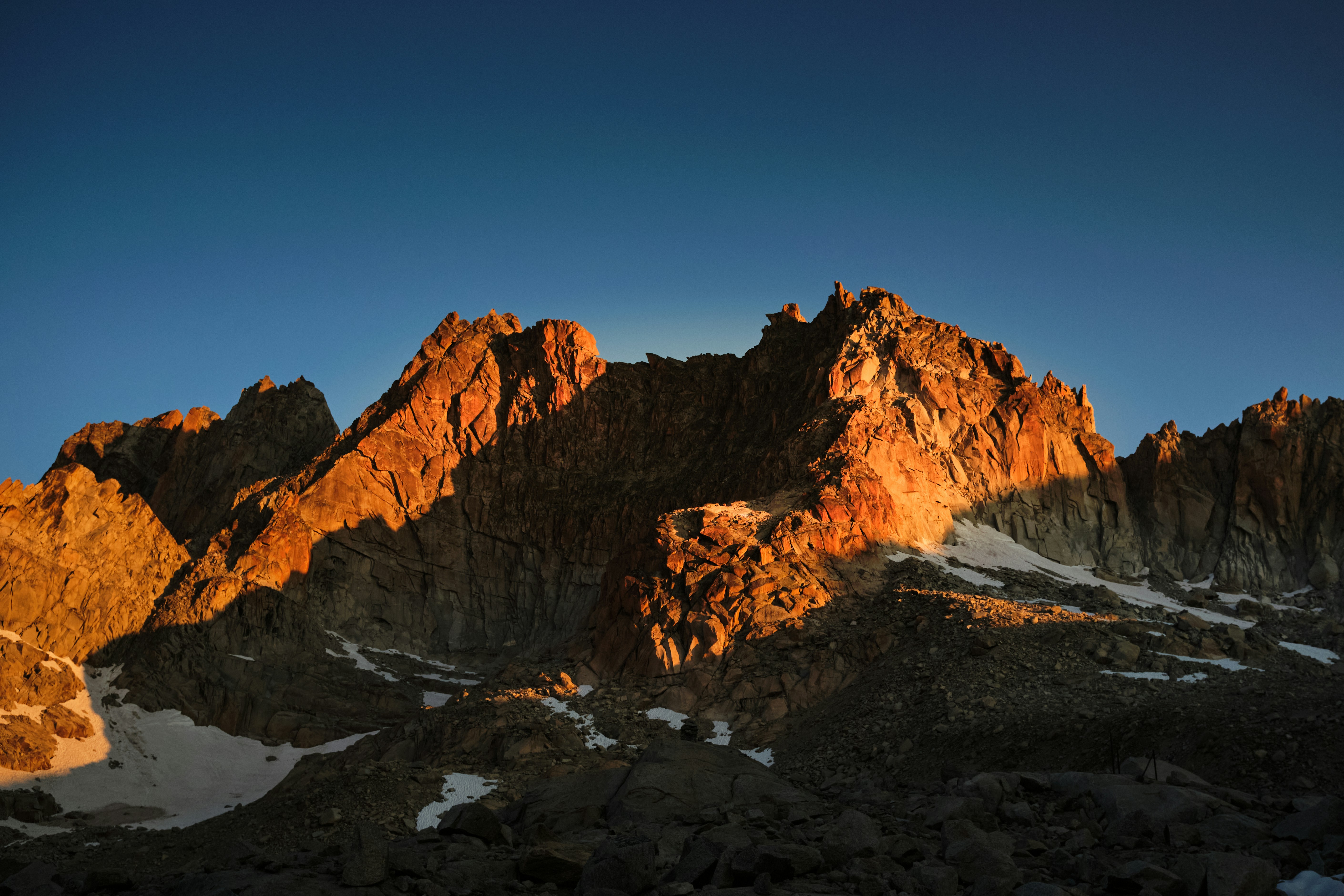 Craggy mountain ridges bathed in warm sunset light against a deep blue sky; rugged rocks with patches of snow define the terrain and depth of the scene.