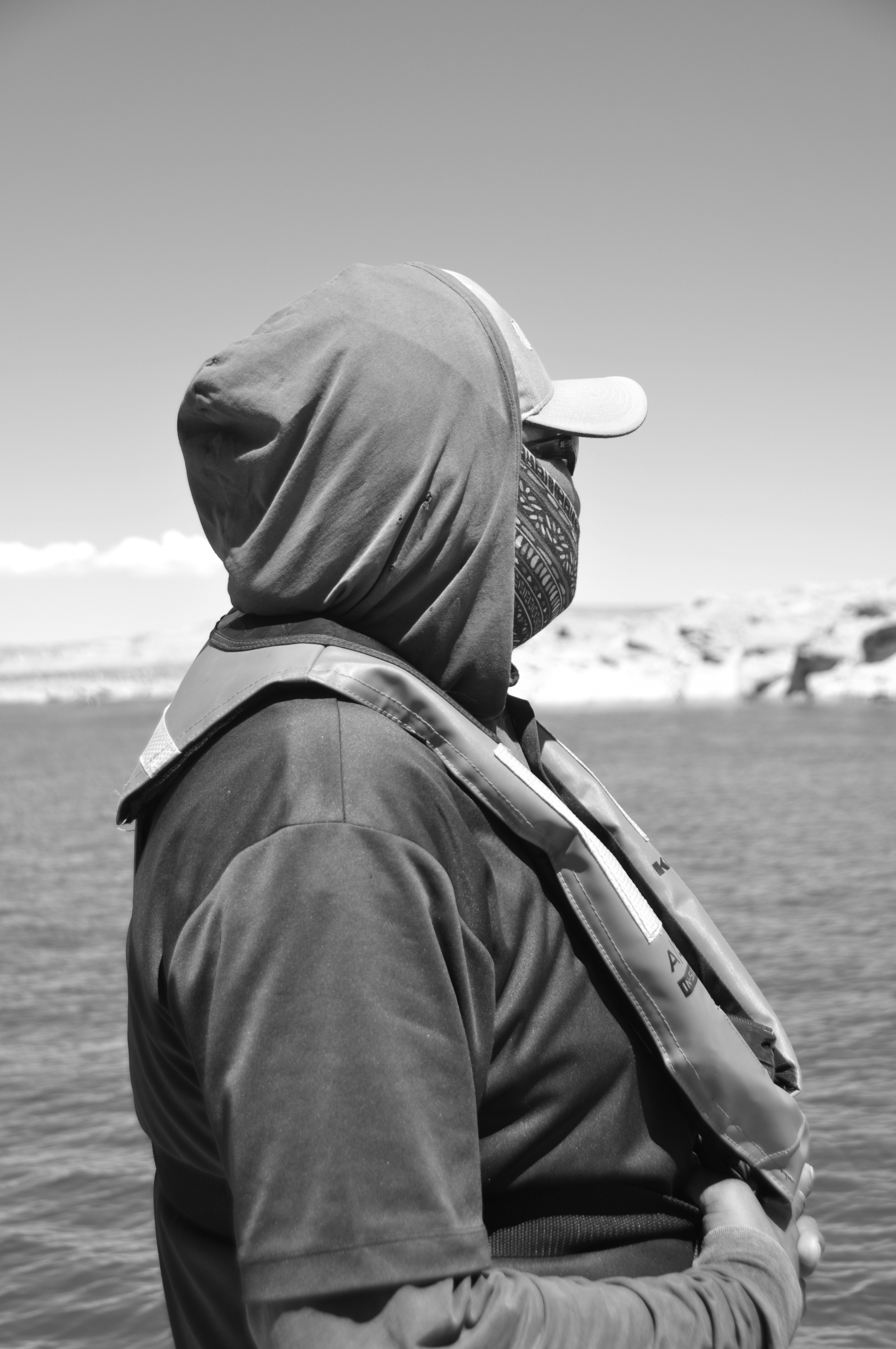 Black-and-white portrait of a person in a hood and scarf facing left, standing at a calm shoreline with distant rocks; a life jacket hints at a maritime context.