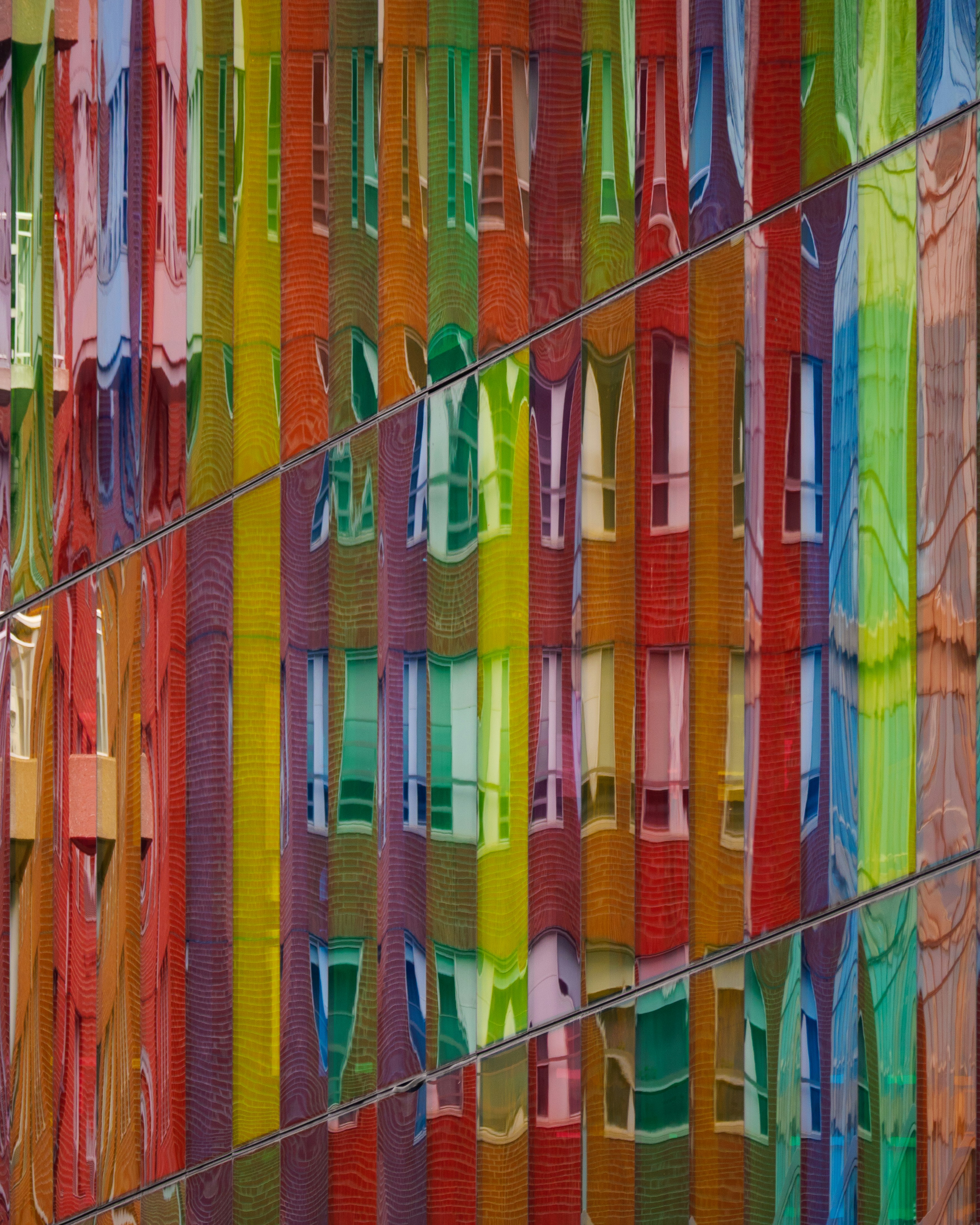 Vibrant multicolored glass panels and diagonal lines form a dynamic, kaleidoscopic facade with distorted window reflections. Abstract architectural art focusing on color and texture.