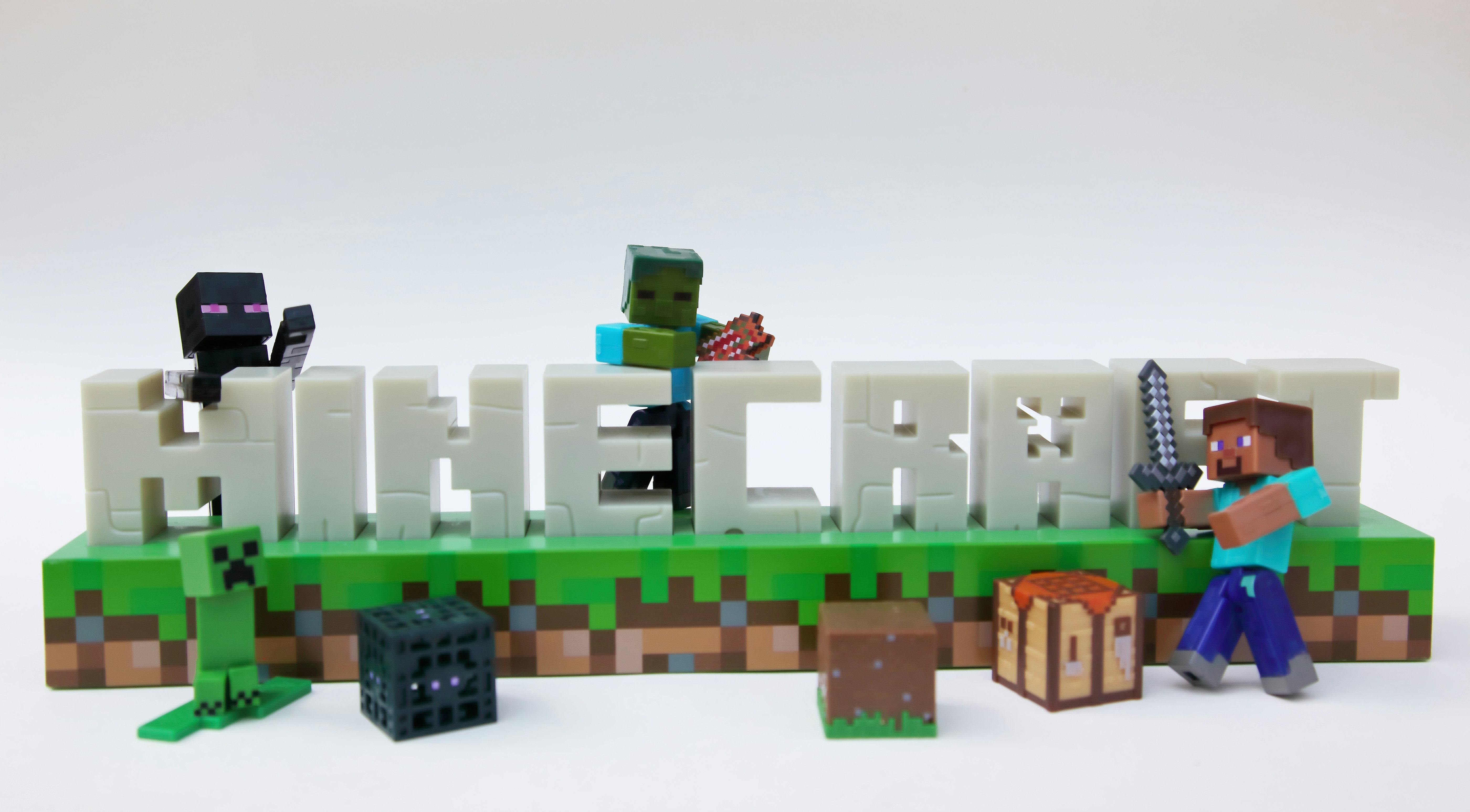 Colorful Minecraft-themed LEGO-like figures and blocks assembled into a playful scene with a raised grass platform and characters; a tiny zombie, creeper, and villagers interact around a chest and crafting table.