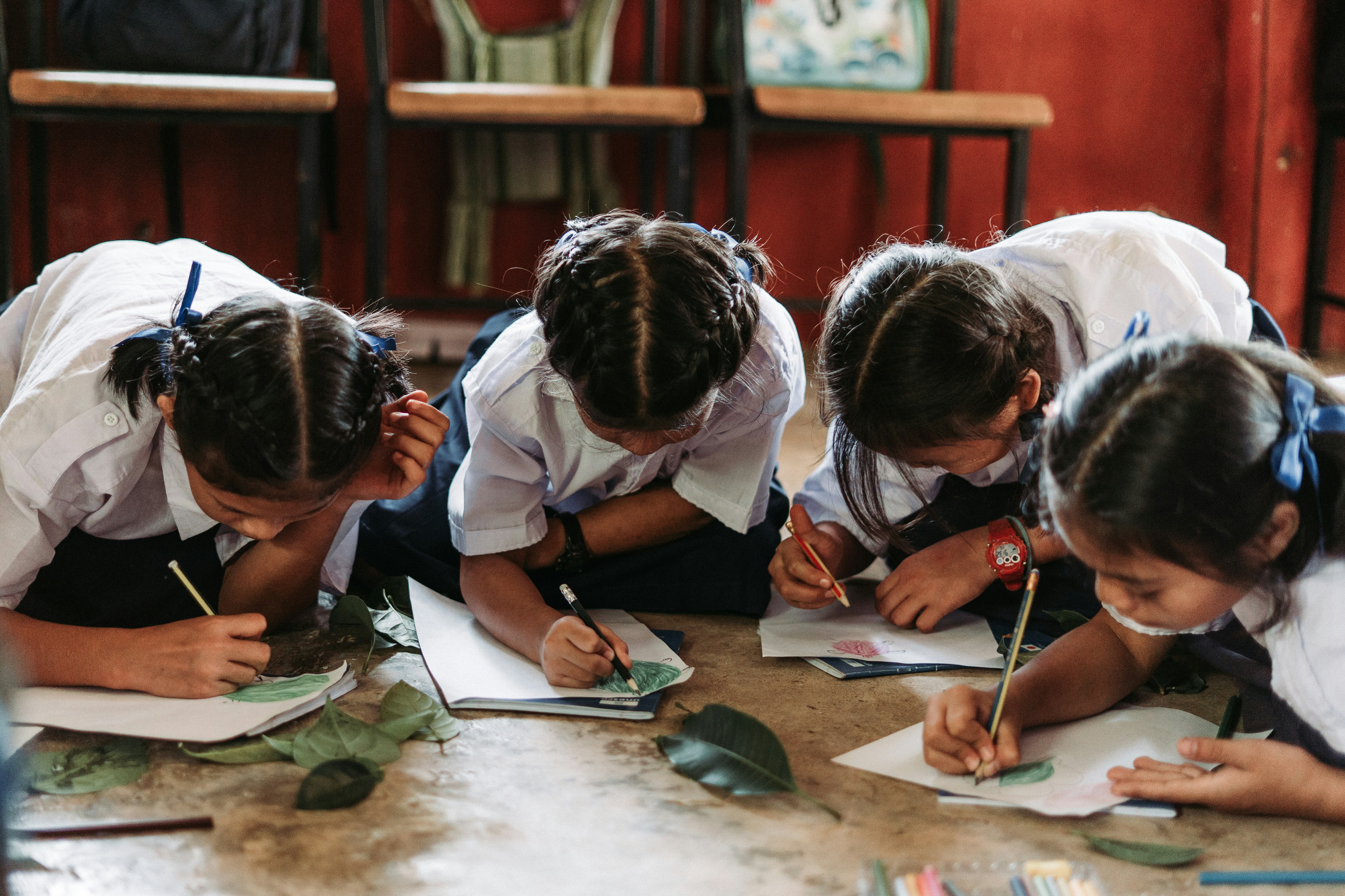 Four young girls in school uniforms sketch on paper, clustered around a low table with leaves and pencils, capturing a moment of shared creativity.
