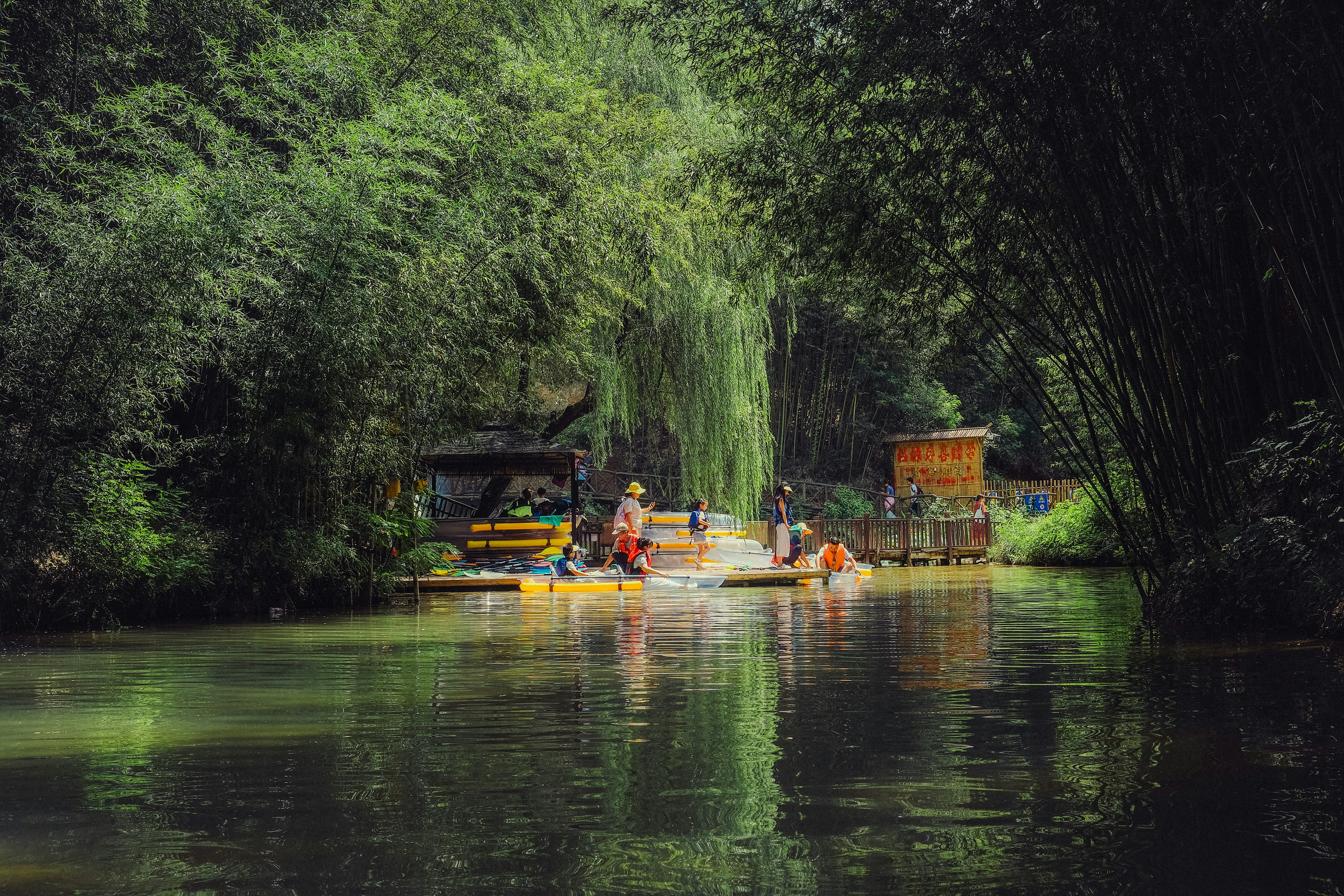 A group of people in orange life jackets gather on a narrow raft-like platform along a calm, green-tinted river bordered by dense trees.