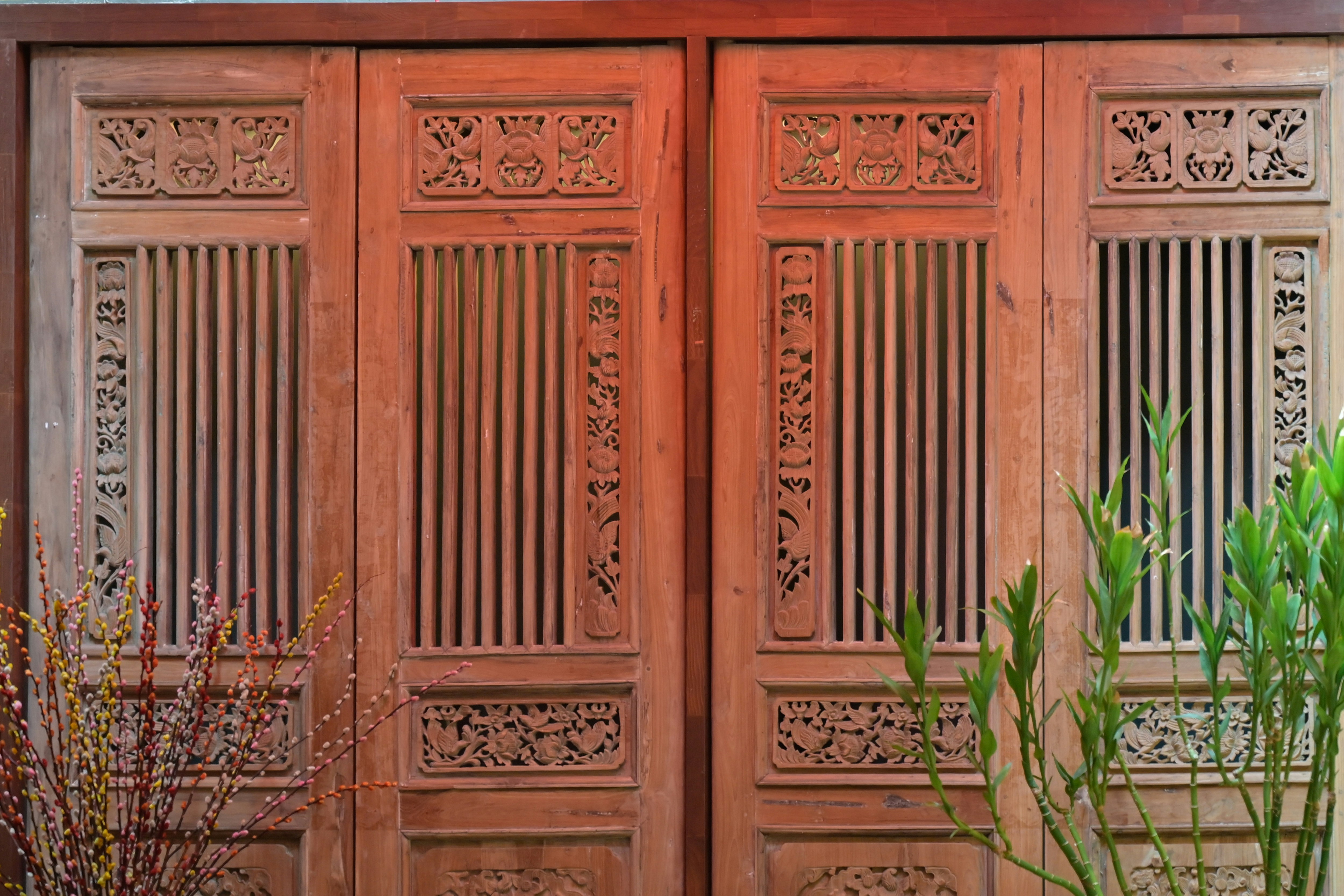 Red carved wooden doors with vertical slats and decorative panels, framed by sparse green shoots in the foreground.