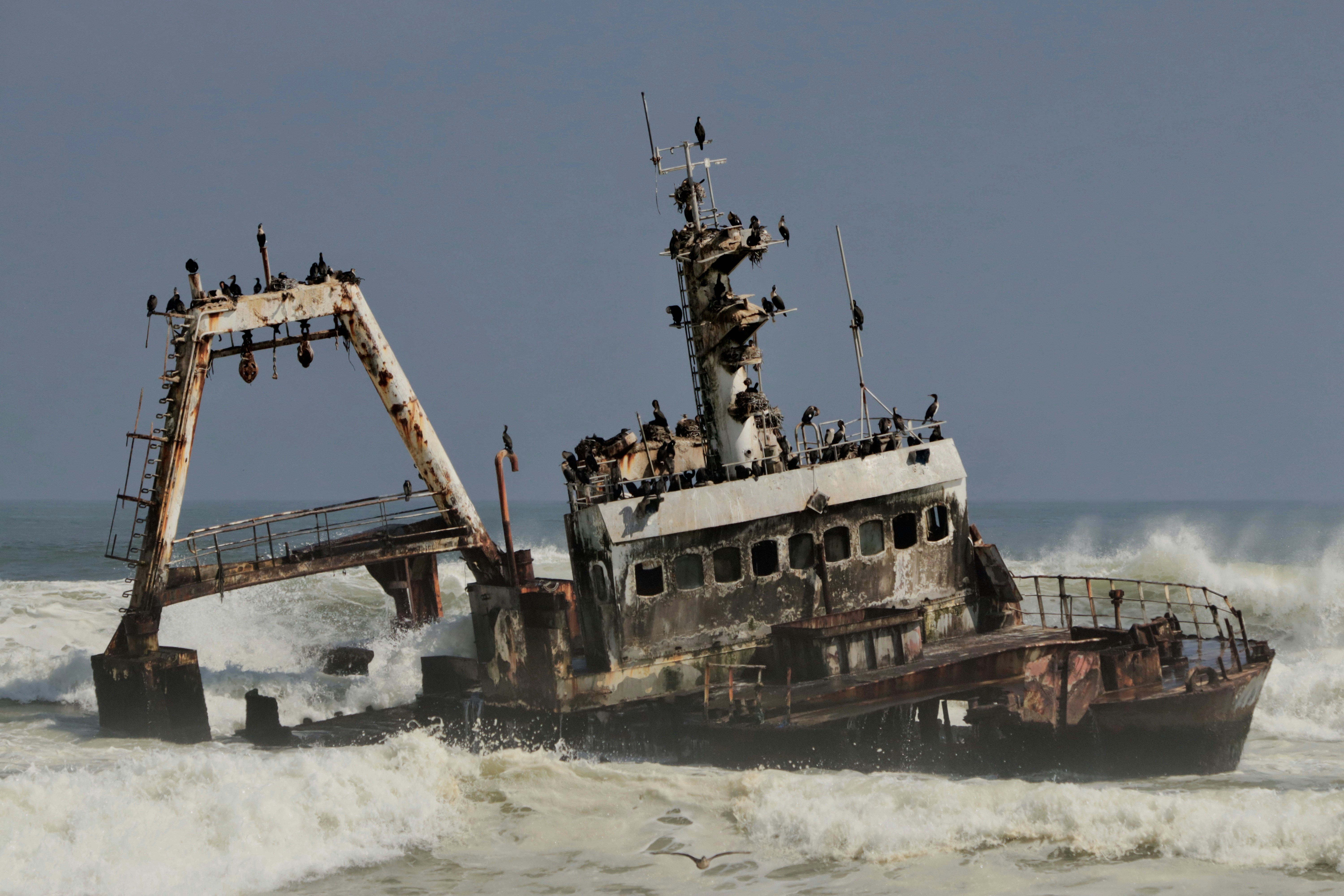 Rusty shipwreck stranded at sea, waves crashing around its battered hull, captured in daylight with a blue sky backdrop.