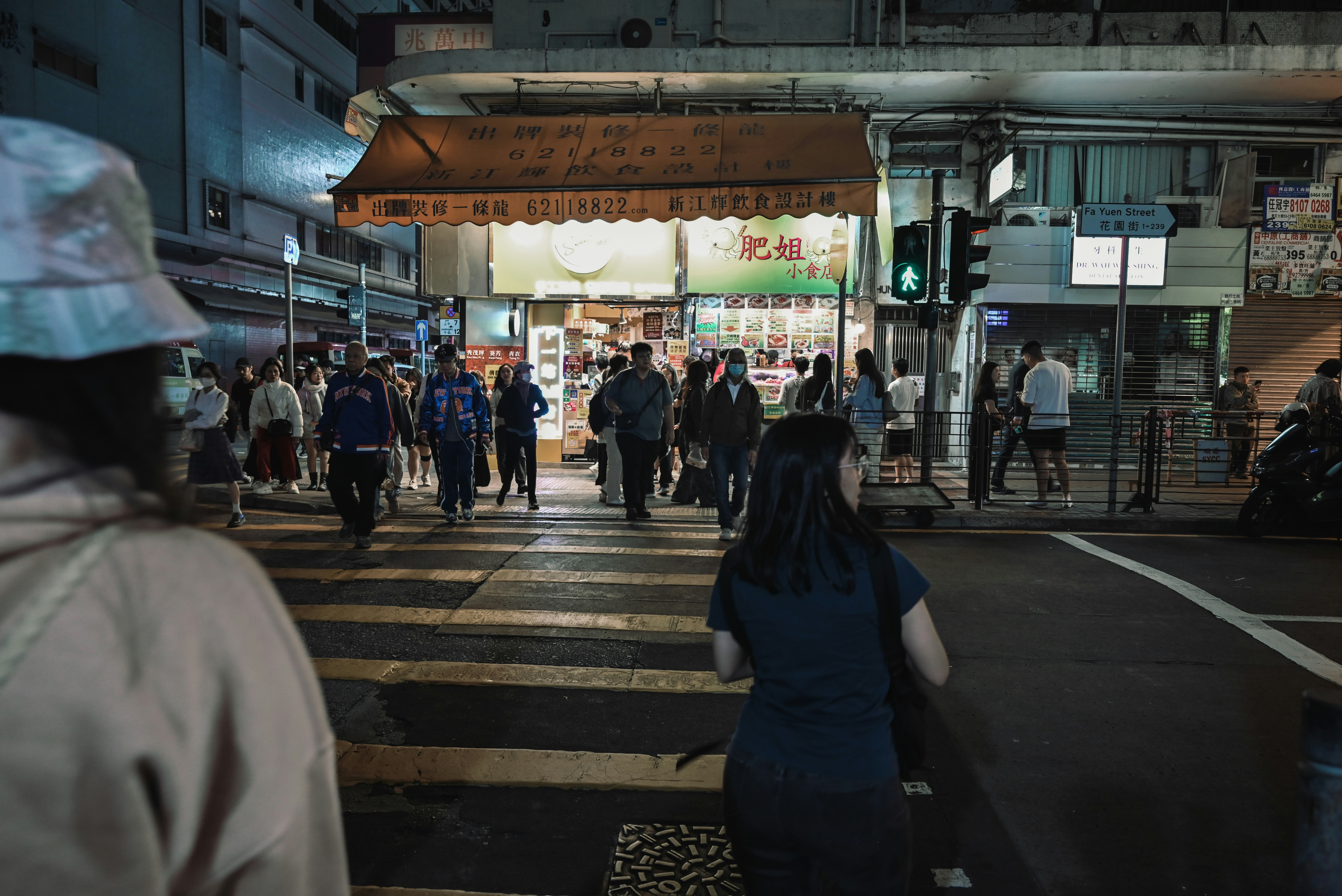 Crowd gathers at a lit storefront doorway as pedestrians cross a city street at dusk; a darkened figure fronts the frame, foreground left, while the shop’s warm glow anchors the background. This is a candid street scene.