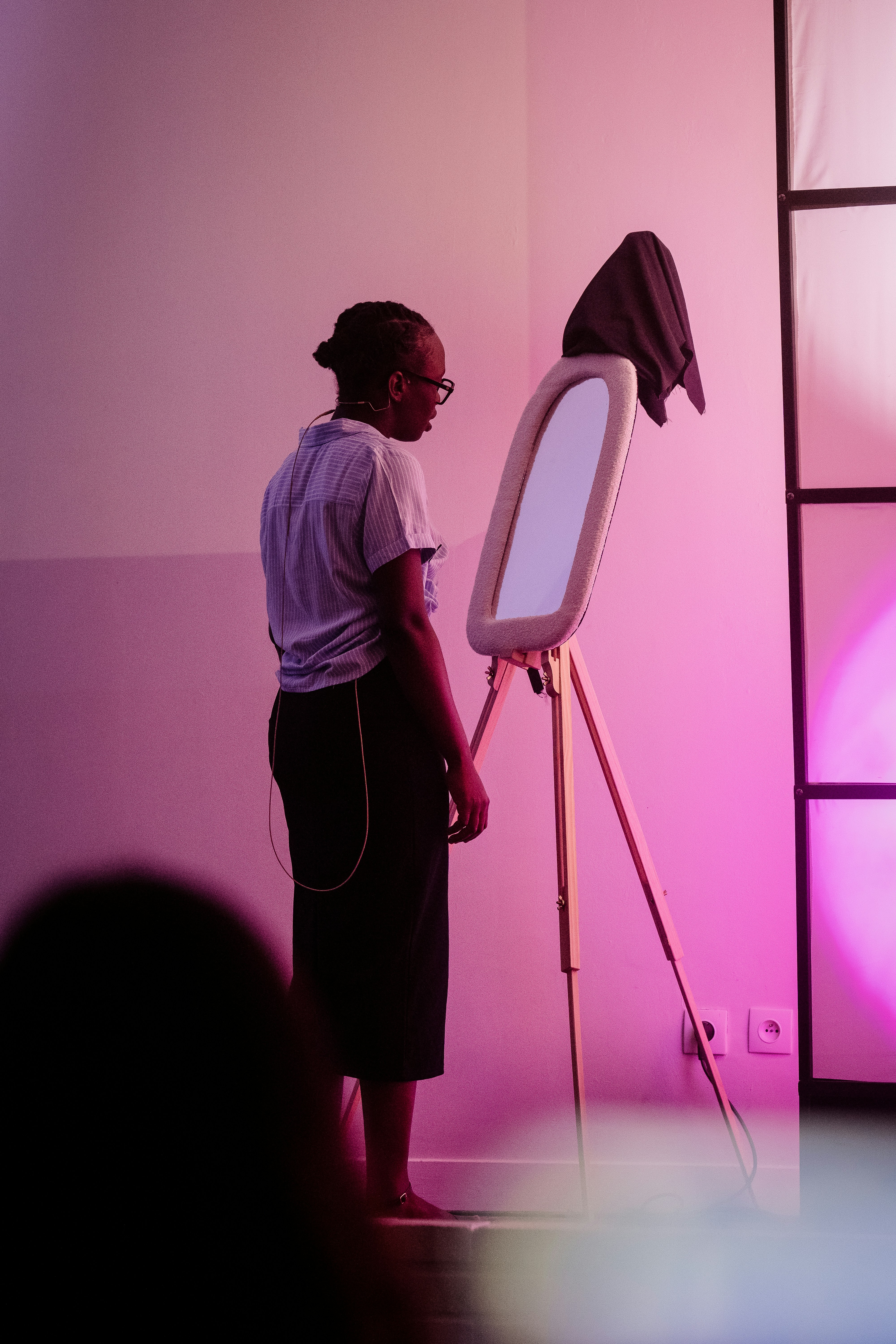 A person stands beside a tall mirror on a tripod, bathed in magenta lighting, with a draped cloth over the mirror and a softly lit backdrop.