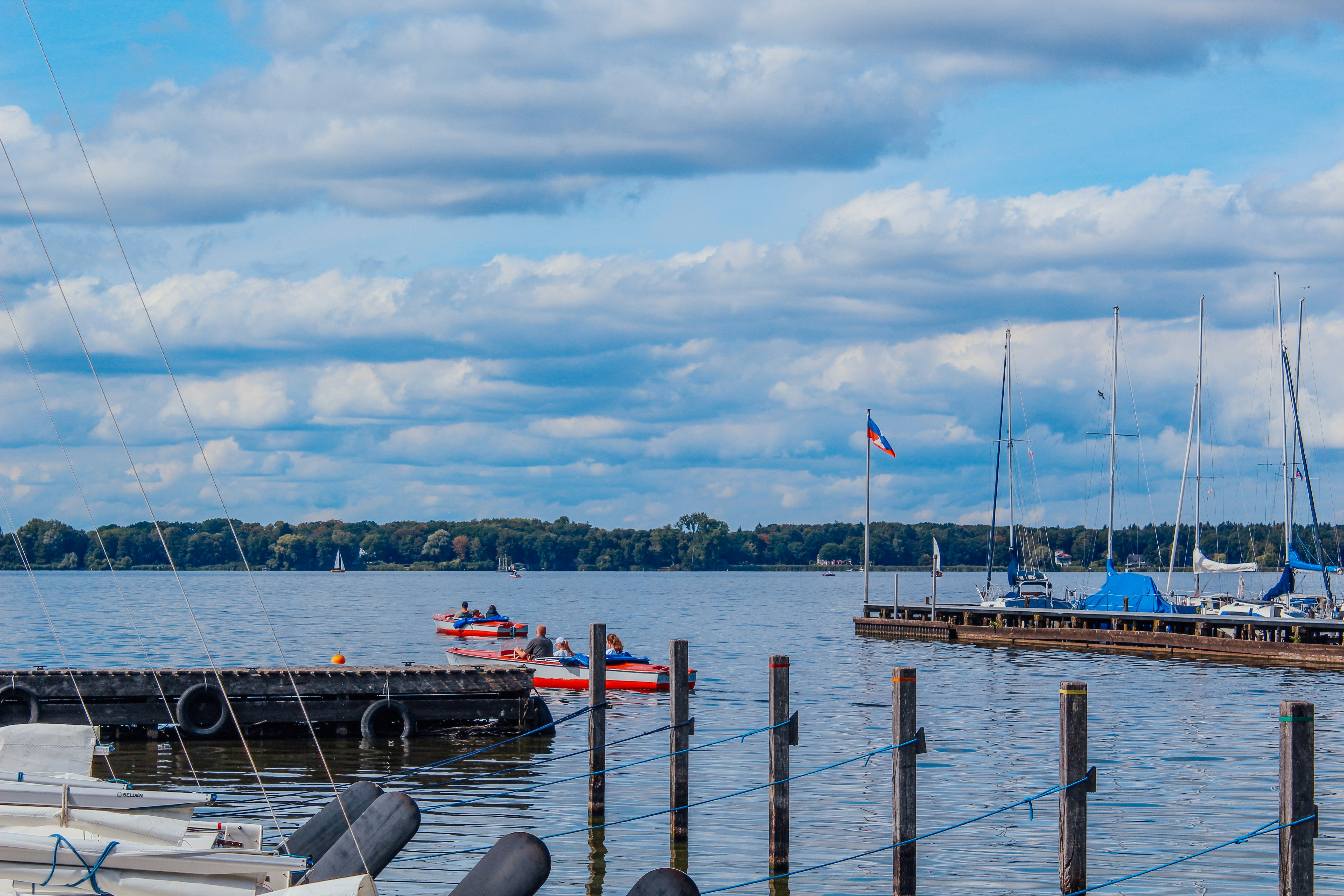 Sunlit marina with boats and sails lined along a calm lake, distant shoreline under a partly cloudy sky.