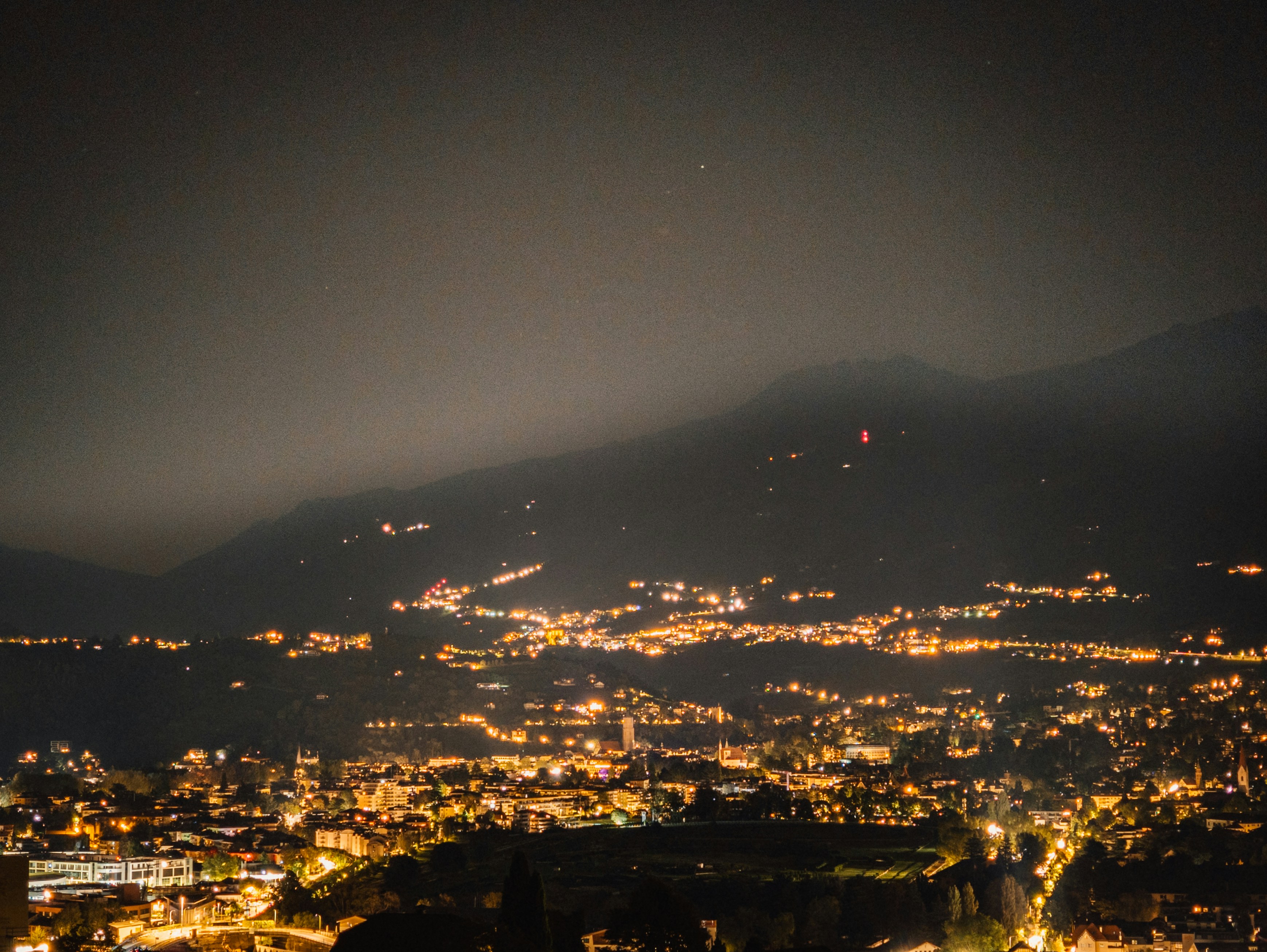 Nighttime cityscape with glowing streets cascading along hillside, captured from a elevated vantage; urban lights and distant mountains form a hazy silhouette.