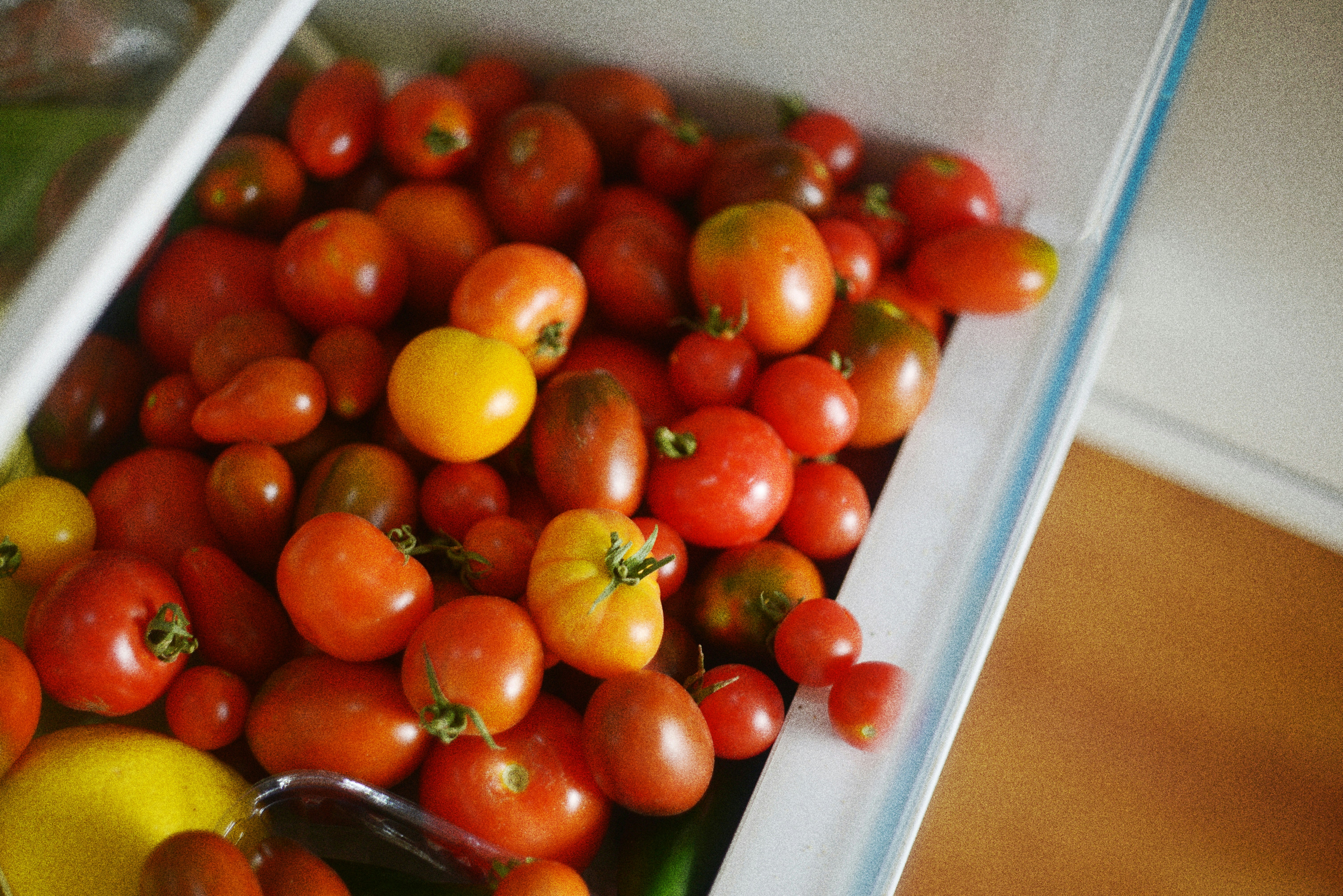 Pile of cherry tomatoes in a shallow plastic crisper tray, with a lime visible at the bottom; a hint of kitchen floor and wall in the background.