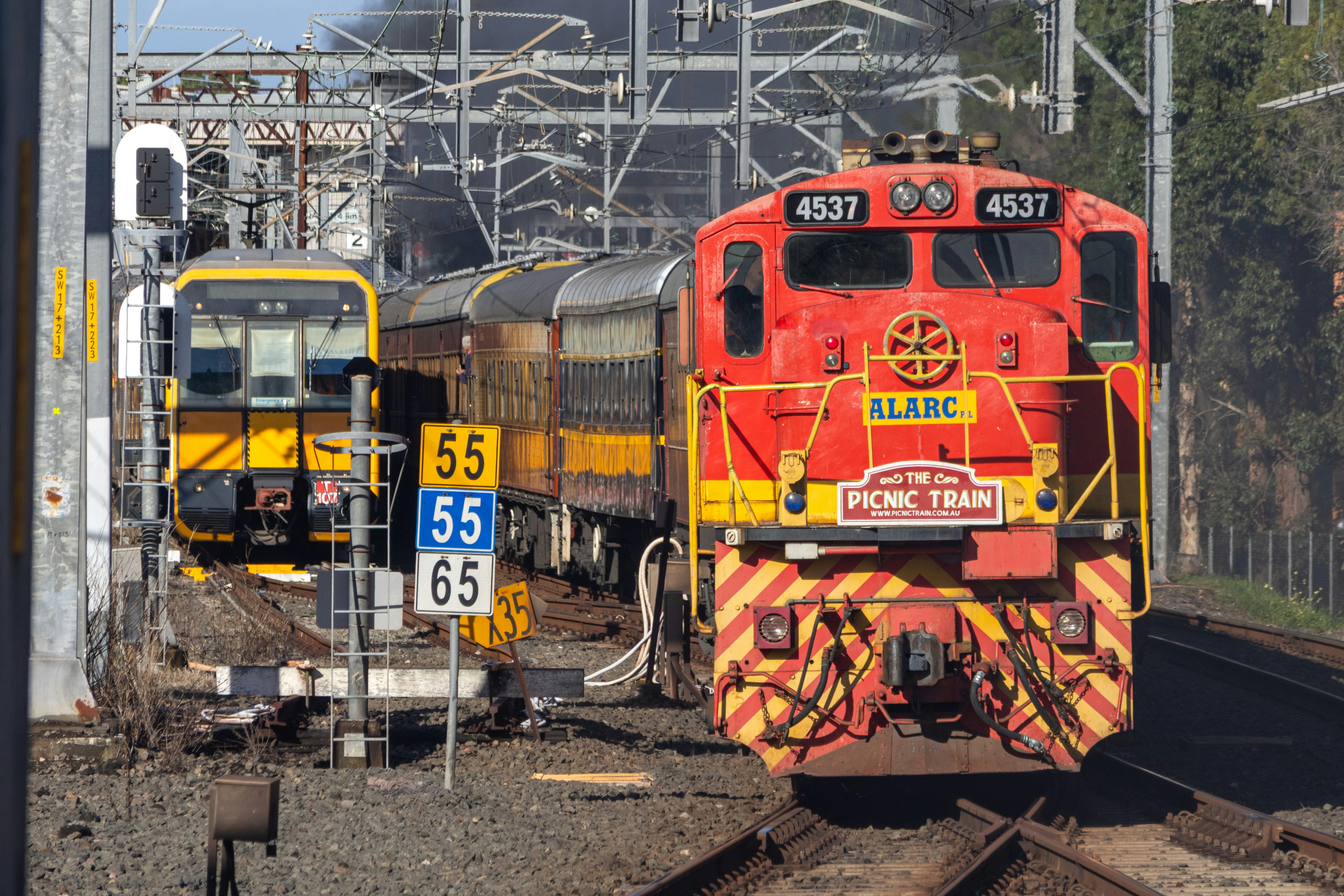 Red locomotive leads a passenger train through a busy yard with a yellow diesel on the left; clear tracks and signaling equipment frame the scene.