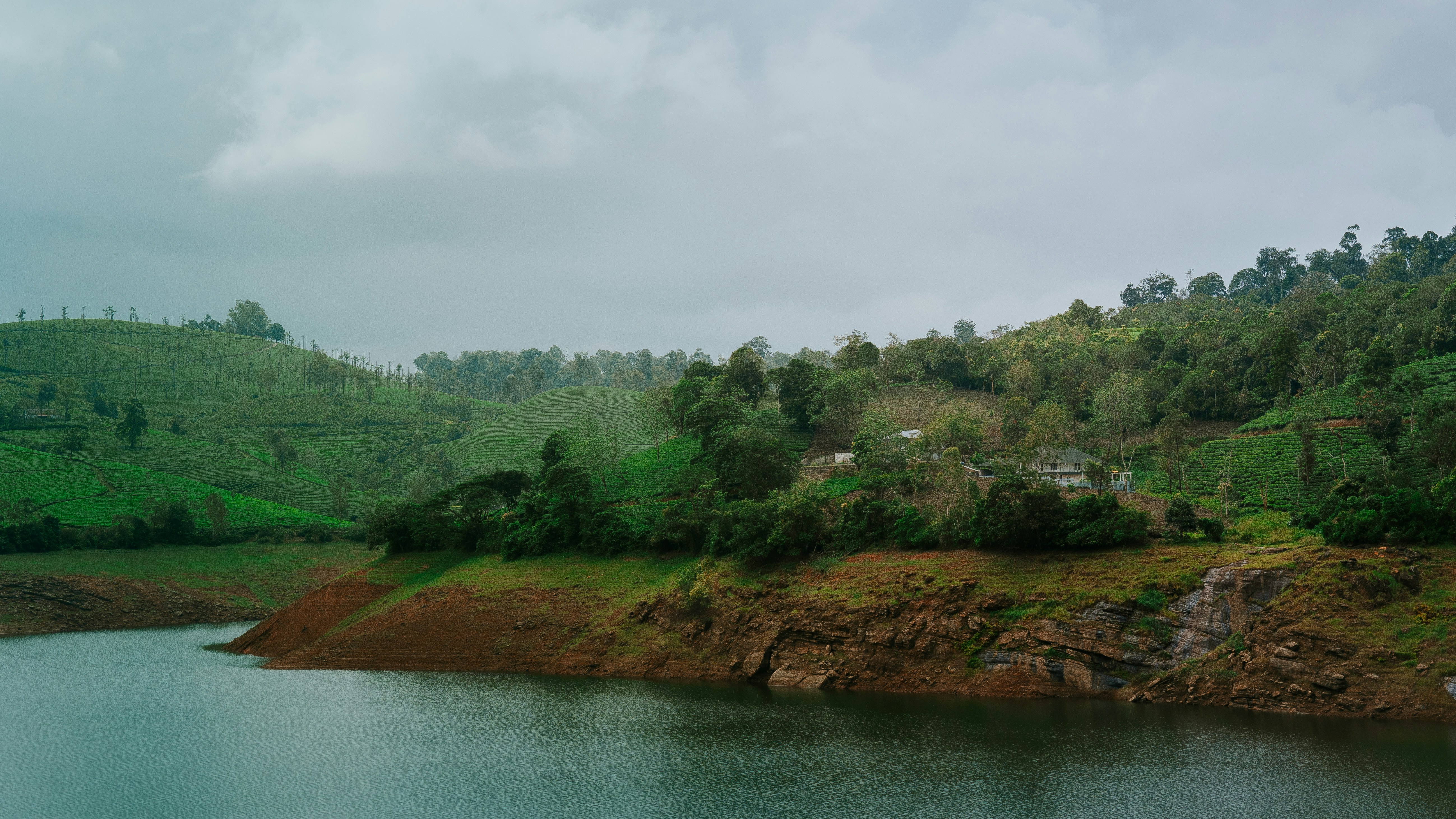 A serene lakeside landscape with rolling green hills and a small settlement along the shoreline. The scene captures calm water, earthy escarpments, and scattered trees under a soft, overcast sky.