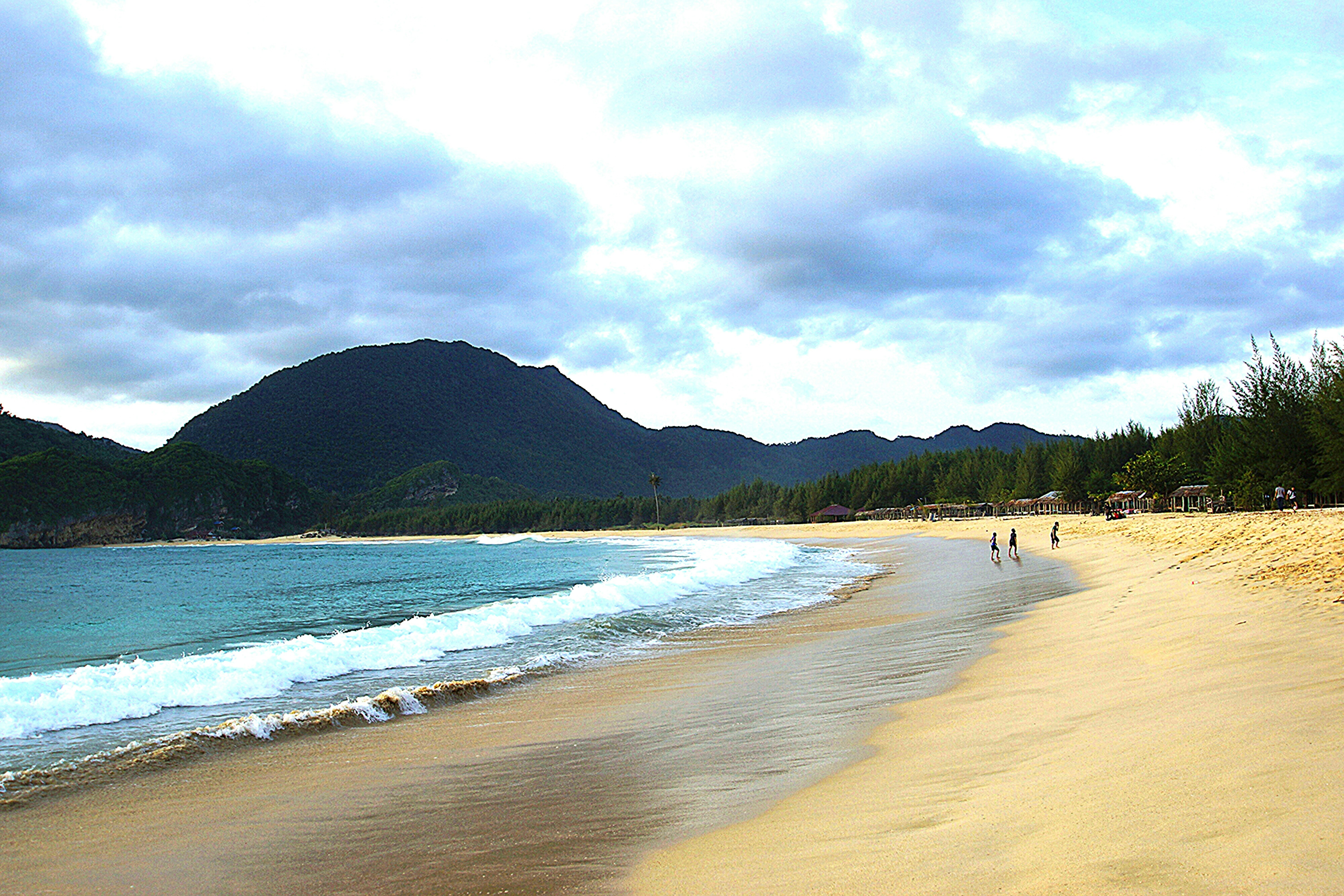 Sunlit golden beach curves along the azure water with distant hills and a few figures walking near the shoreline. Illustration-like balance and soft clouds.
